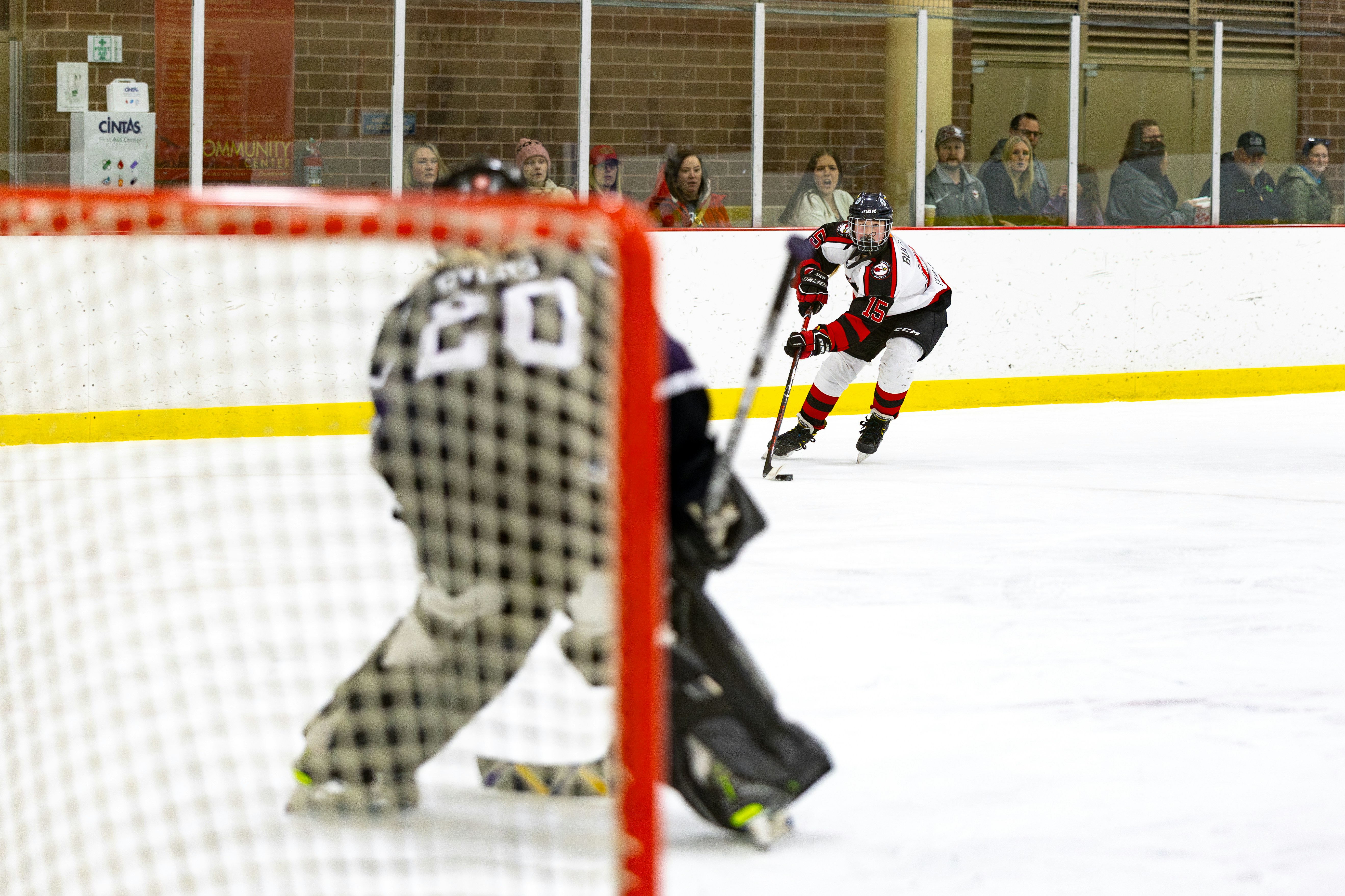 Hockey photograph capturing a white-jersey skater advancing toward the net as the goalie closes in on defense. Spectators are visible along the boards in the background.