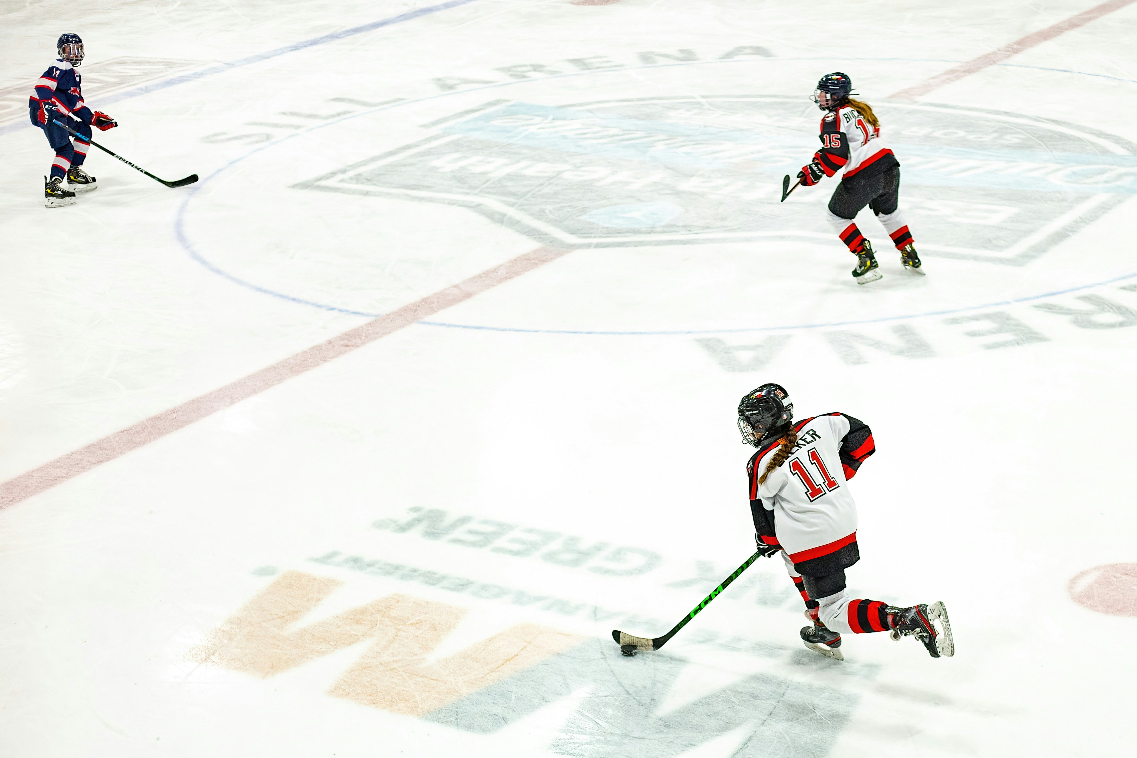 Two young hockey players compete on the ice, with one skating towards the puck while the other maintains defensive positioning. The arena's branding is subtly visible beneath the ice.
