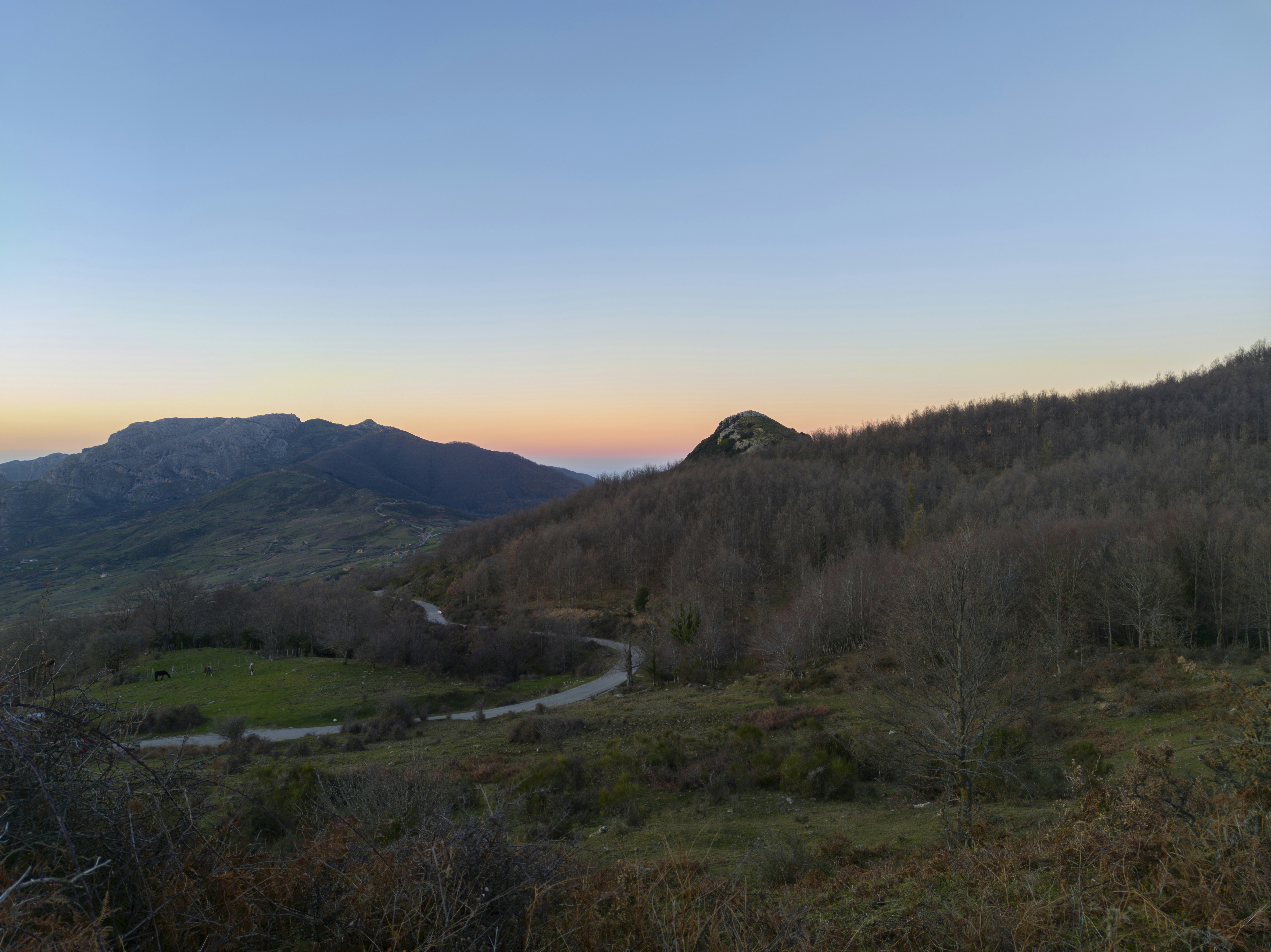 Winding road through hilly landscape at dusk, with a fading sunset over distant mountains.