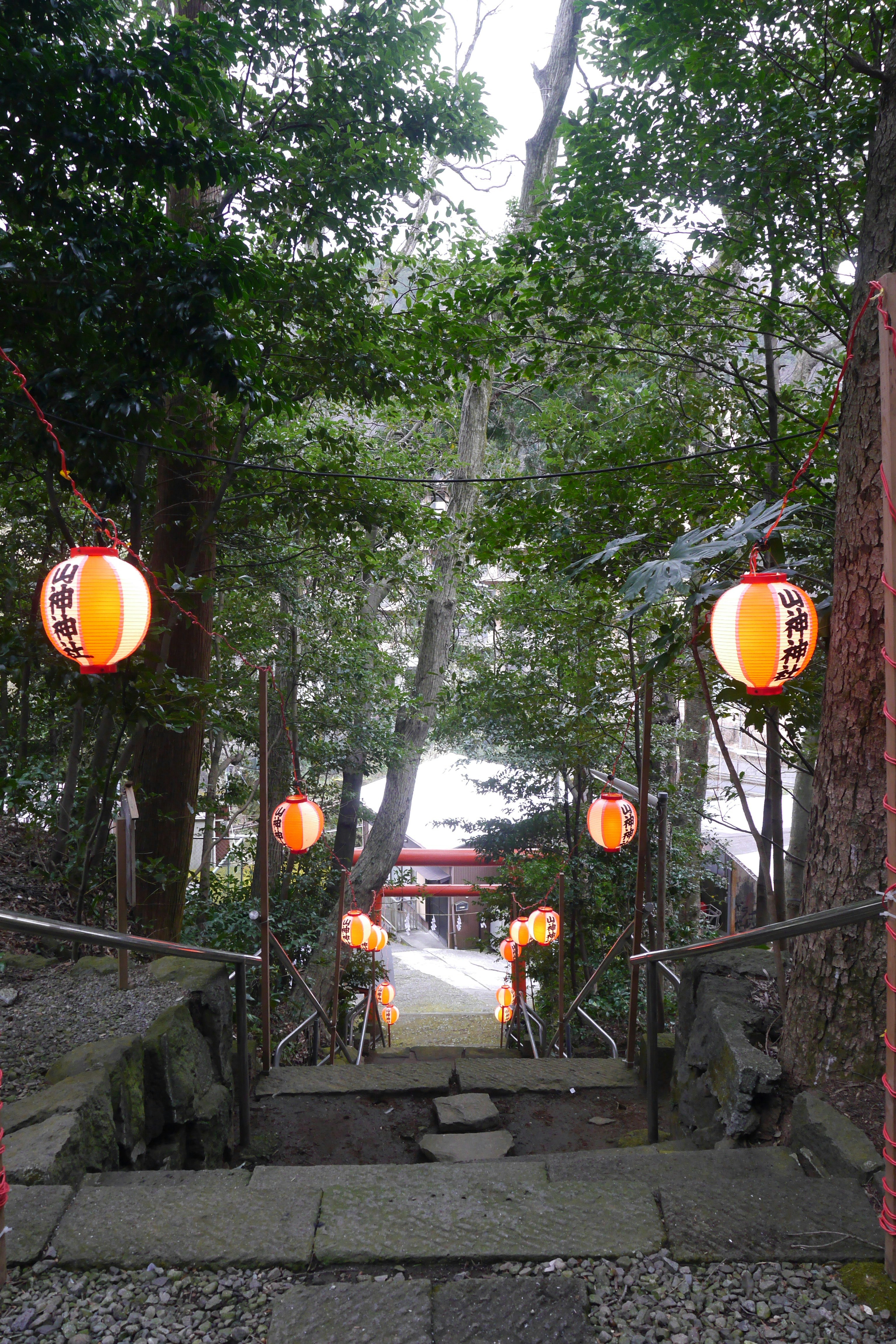 Stone steps descend through a shaded shrine path lined with orange lanterns, converging toward a distant torii gate.