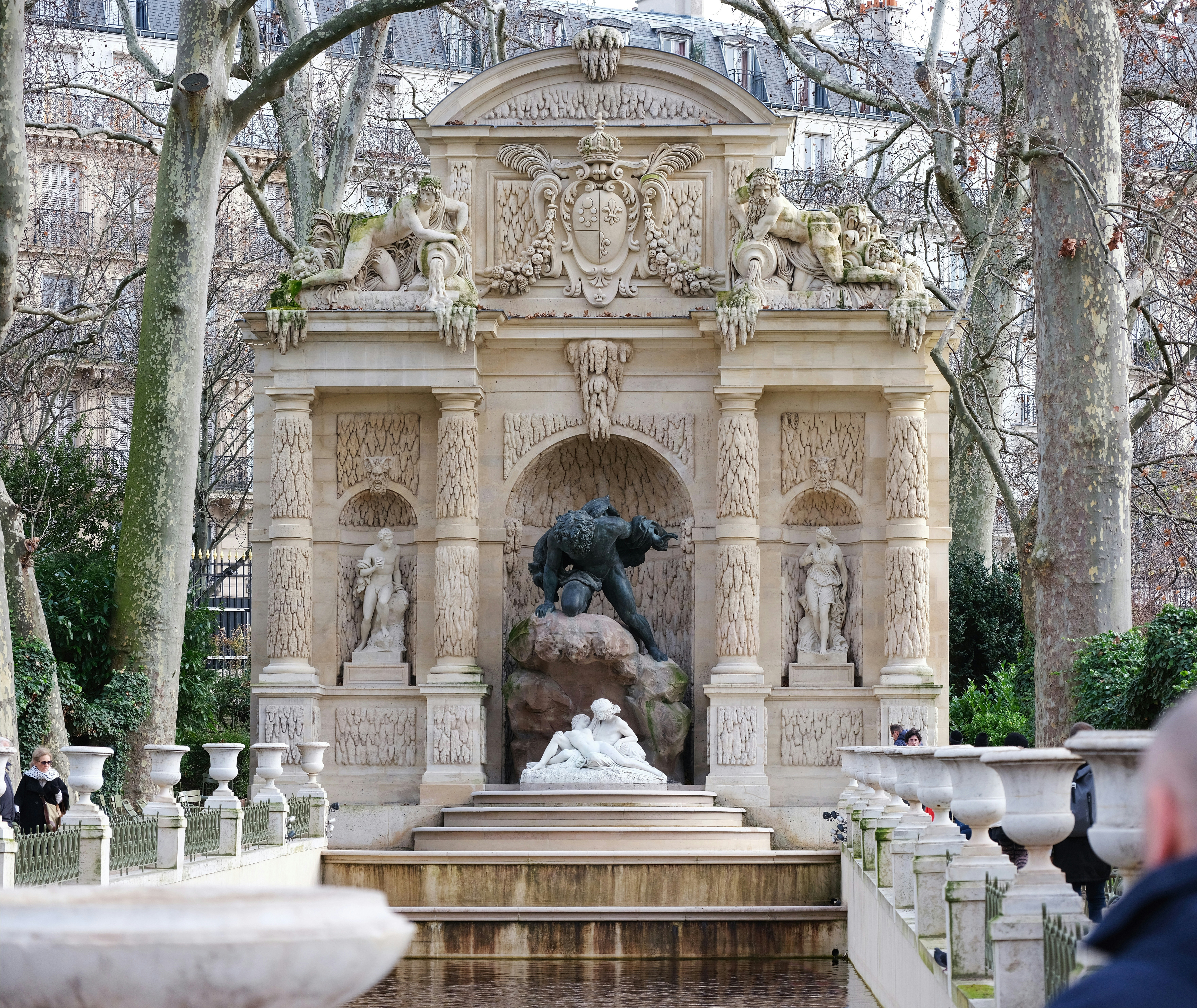 Intricate stone architecture of the Medici Fountain framed by bare winter trees in a Paris park.