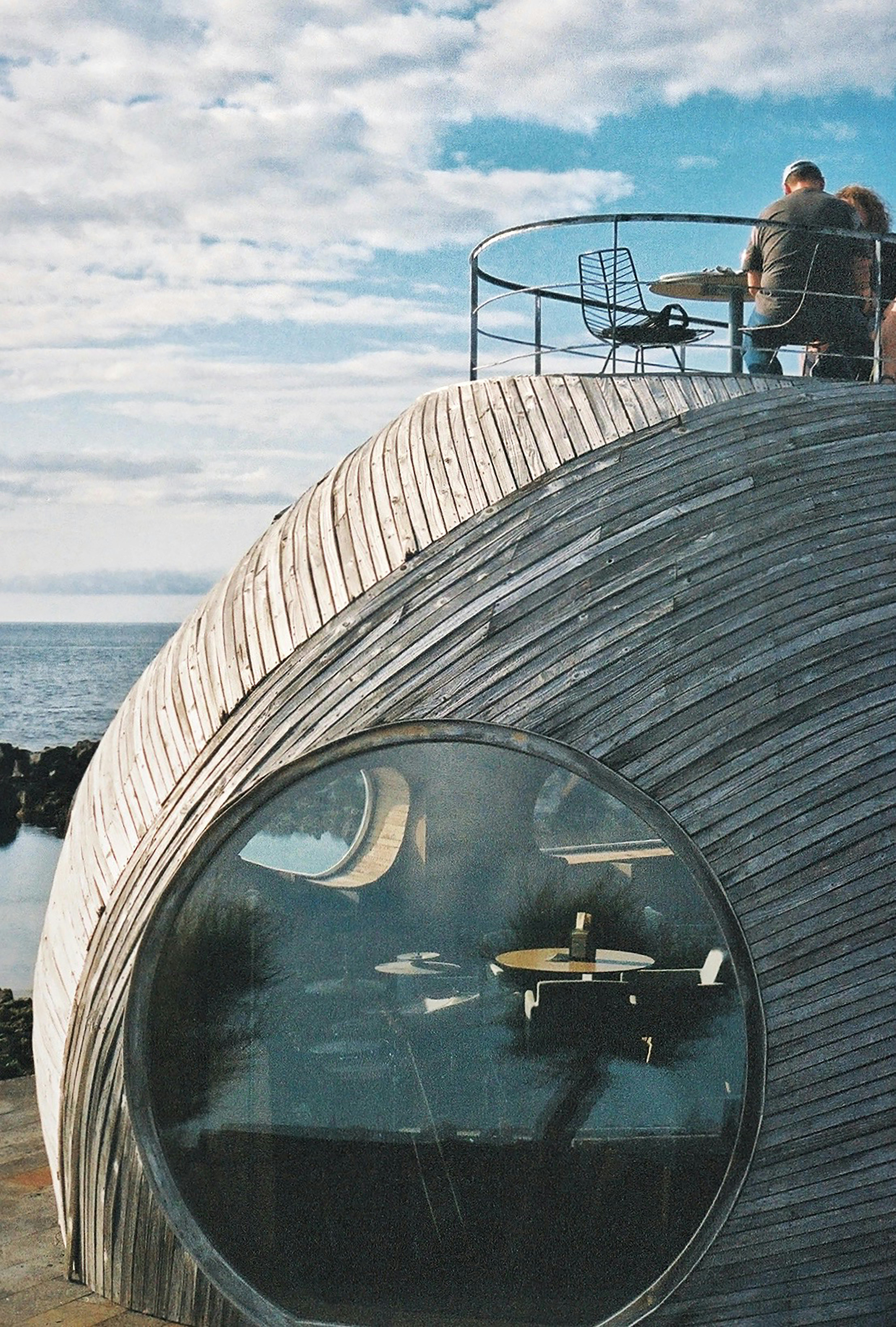 Rounded wooden dome with a prominent circular glass window sits by the coast, its interior dining area faintly visible. Two people stand at a railing atop the structure, gazing toward the sea on a bright, partly cloudy day.