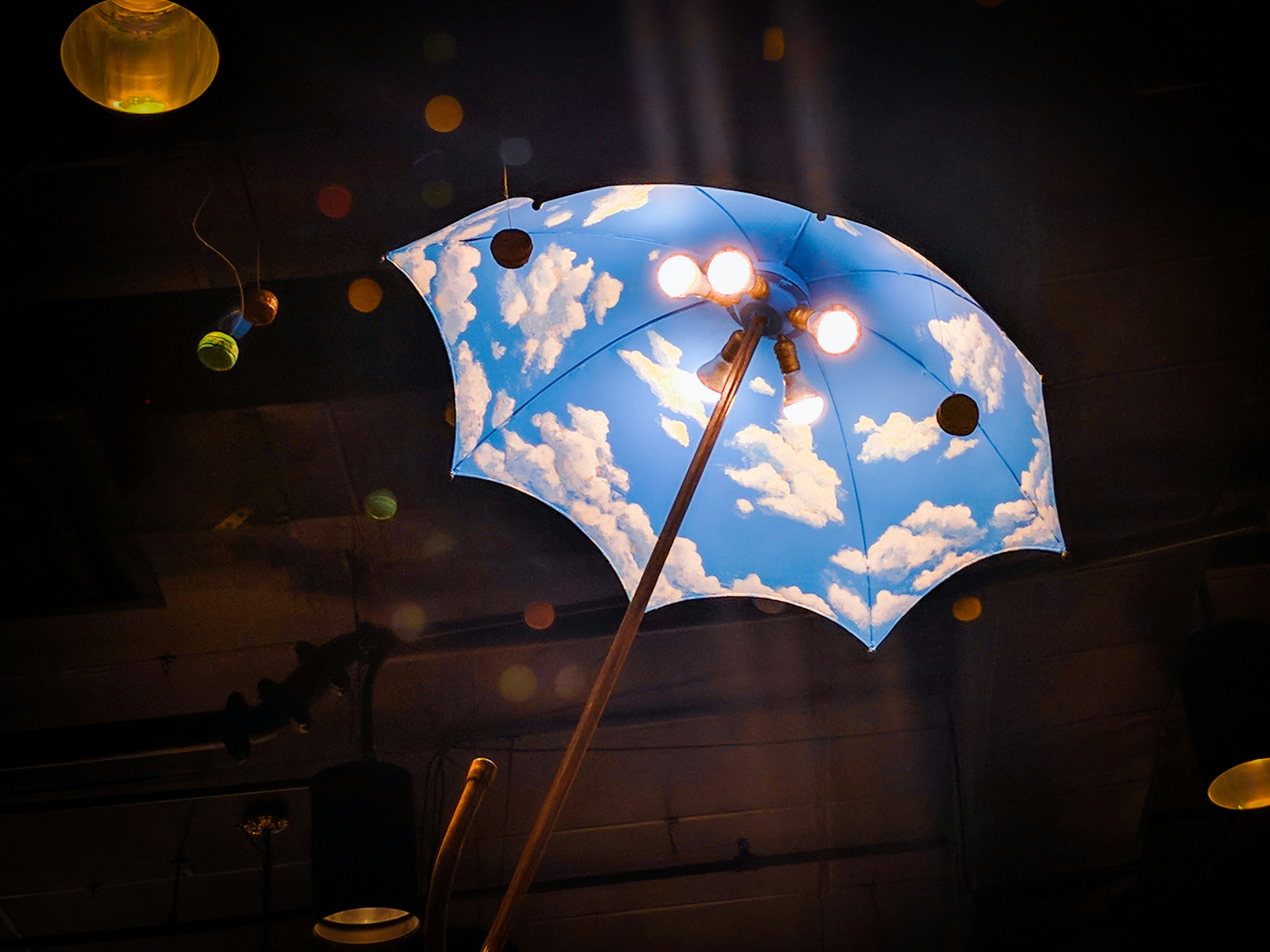 Blue umbrella with cloud patterns lit from beneath by warm lights in a dimly lit space.