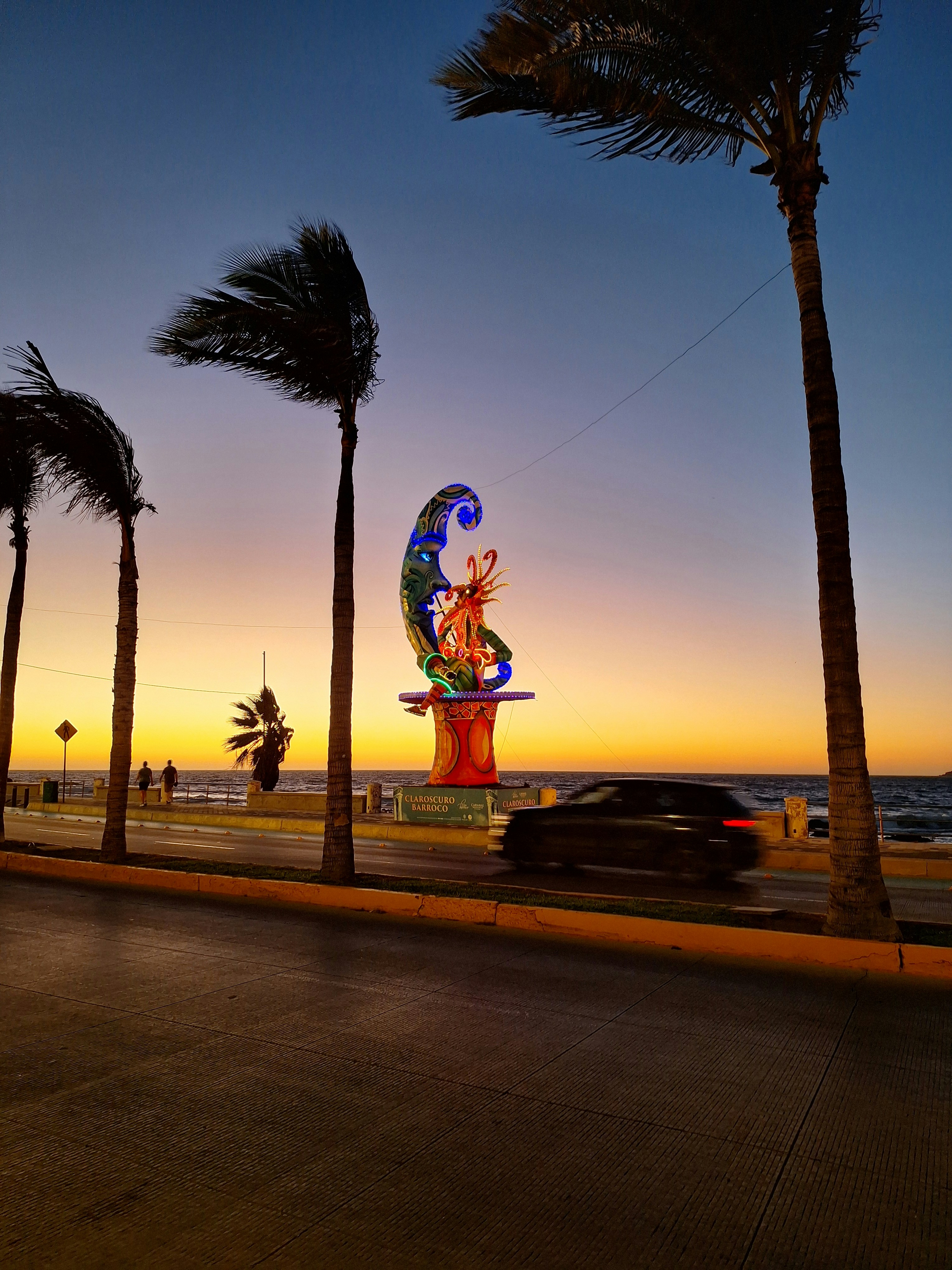 Colorful sculpture illuminated against a vibrant sunset, framed by swaying palm trees along the shoreline.