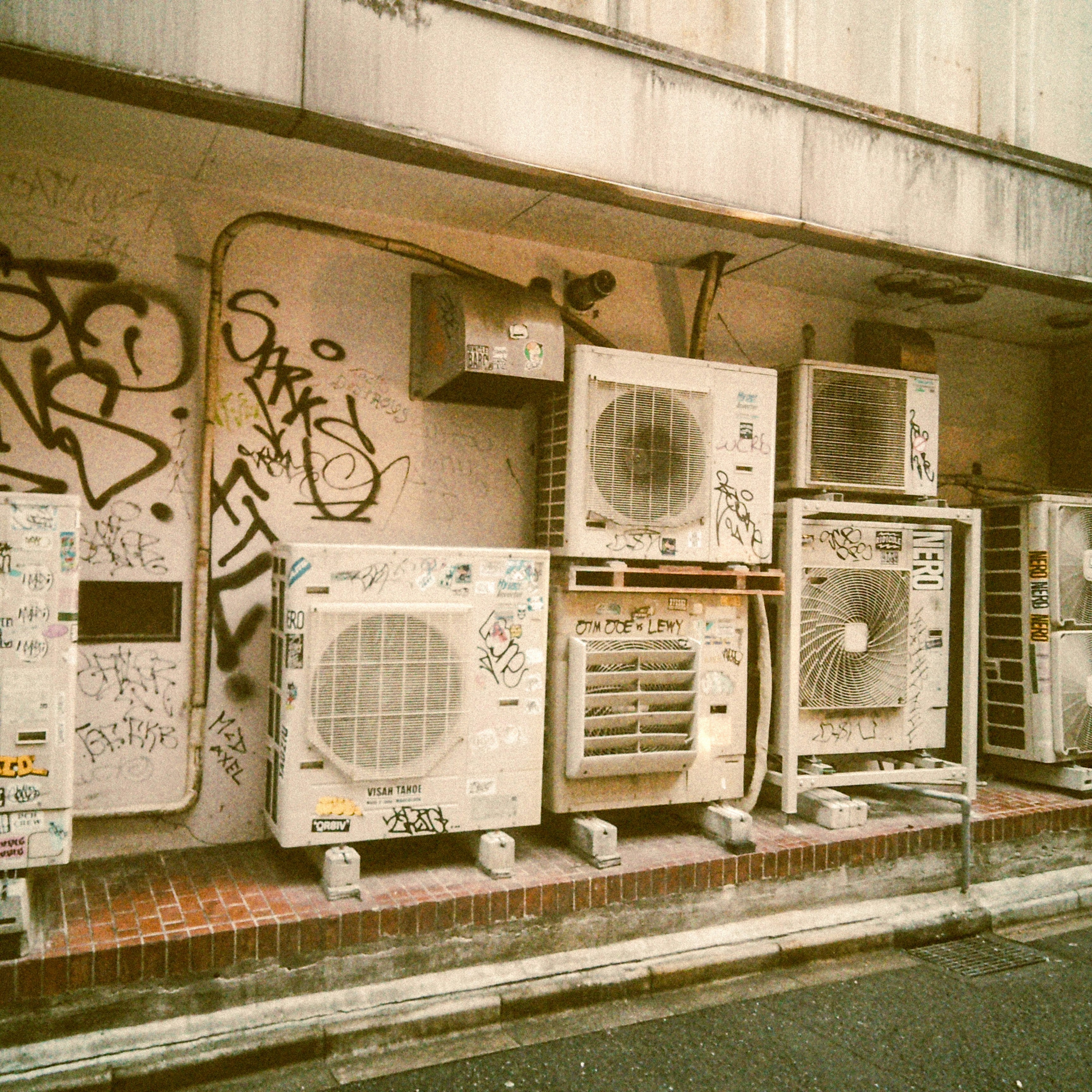 Row of outdoor air conditioning units along a graffiti-splashed wall in an urban setting; weathered surfaces and exposed wiring create a gritty, industrial mood.