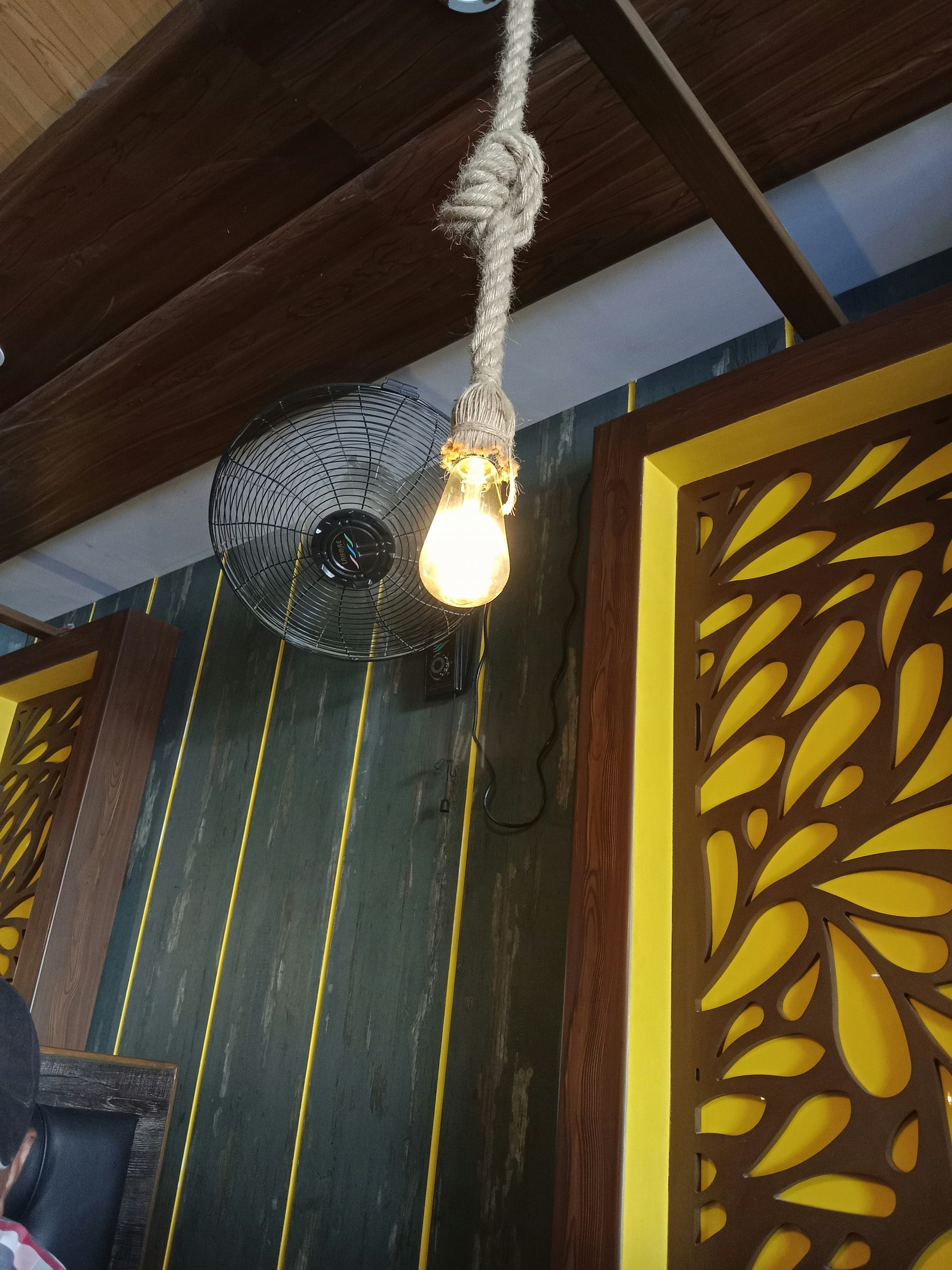 Warm Edison bulb hangs from a knotted rope beside a wall fan and carved yellow panels, highlighting a cozy, decor-forward interior.