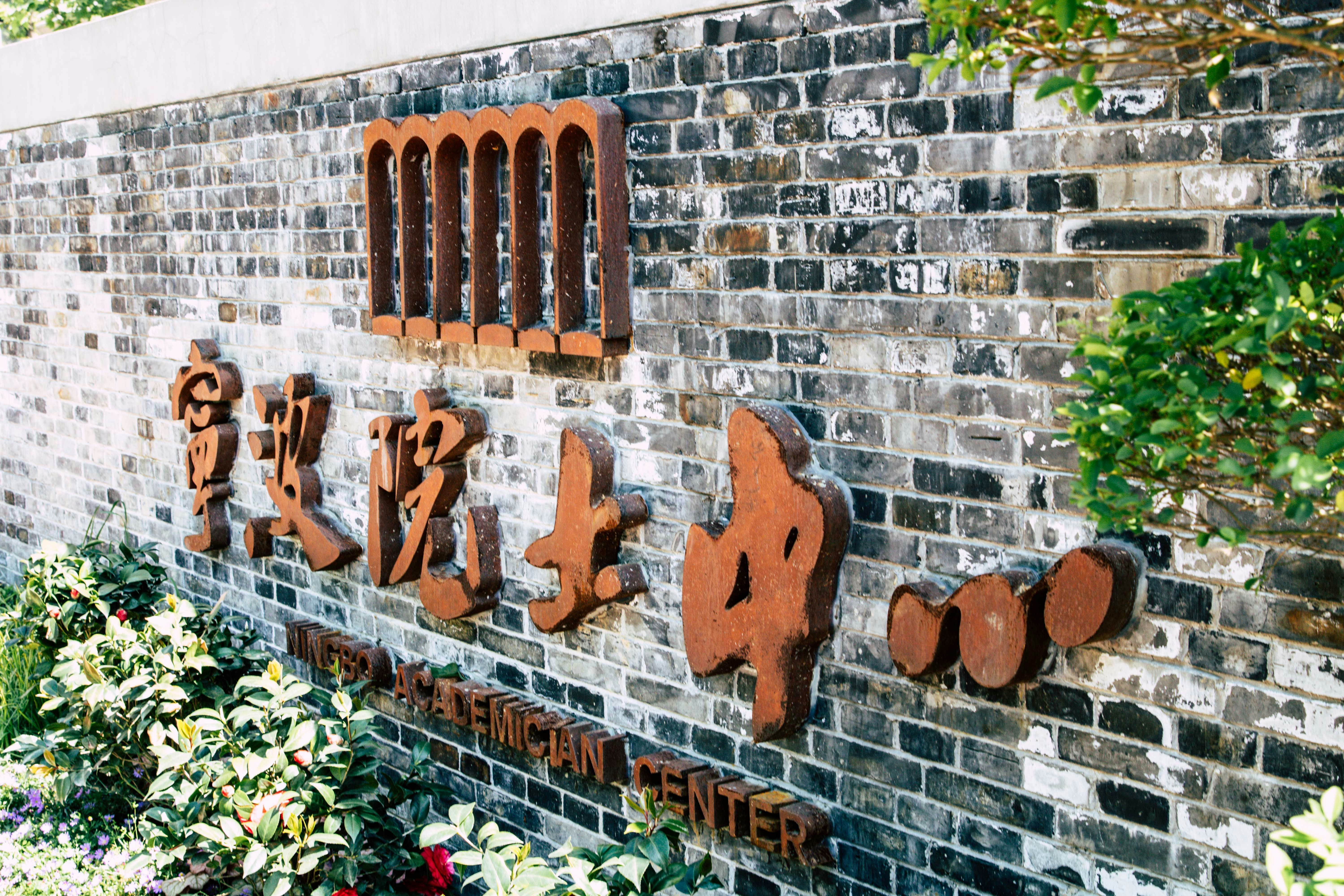 Rustic signage of a cultural center, featuring intricate lettering against a textured brick background, surrounded by vibrant greenery.
