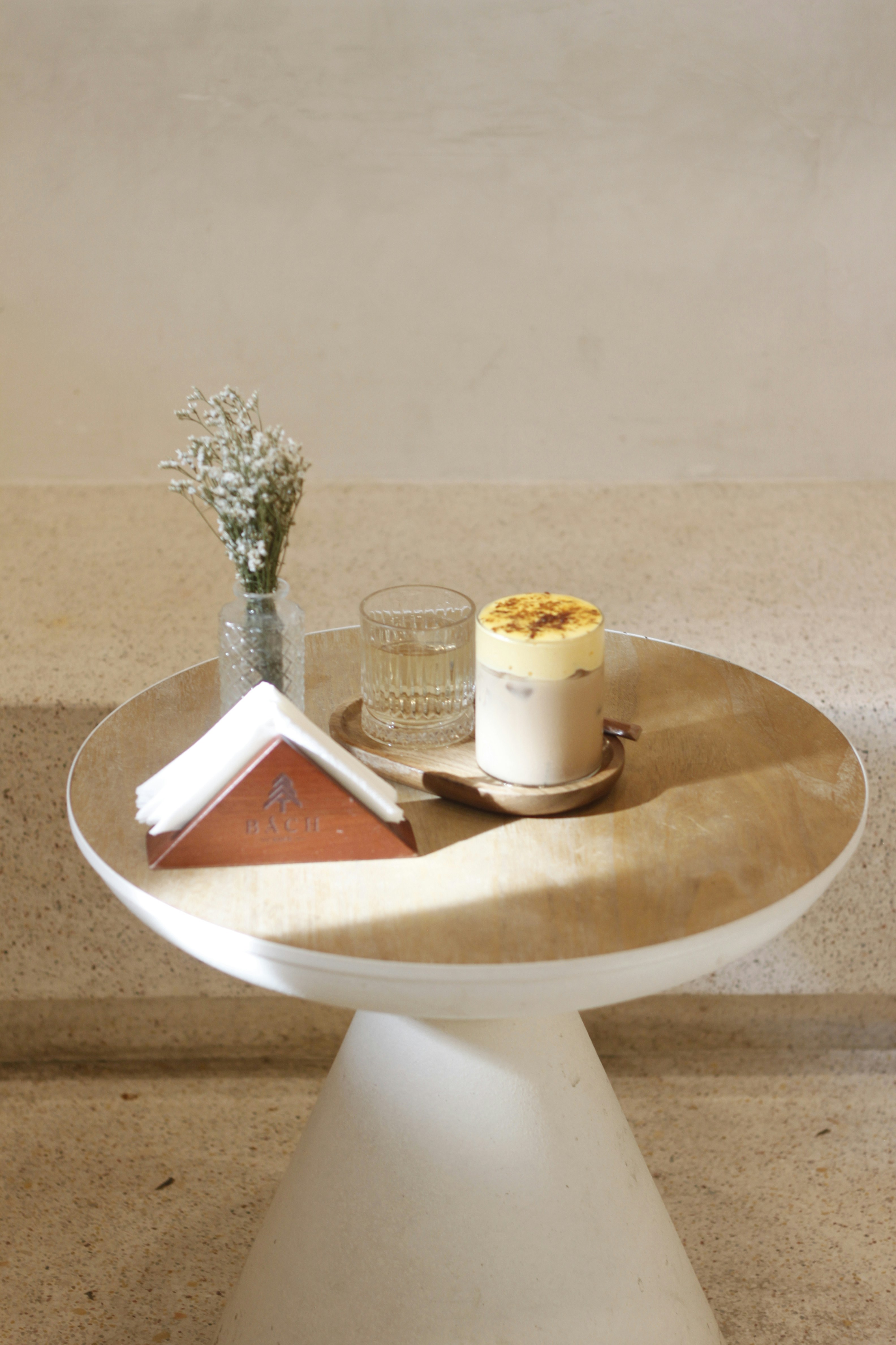 A serene café scene featuring a small round table with a rustic wooden top, adorned with a glass of iced coffee topped with foam, a clear glass of water, and a small vase of delicate white flowers. Warm sunlight casts soft shadows across the table, enhancing the muted beige and cream tones of the setting. The image exudes a calm and inviting atmosphere, perfect for a quiet moment of relaxation.