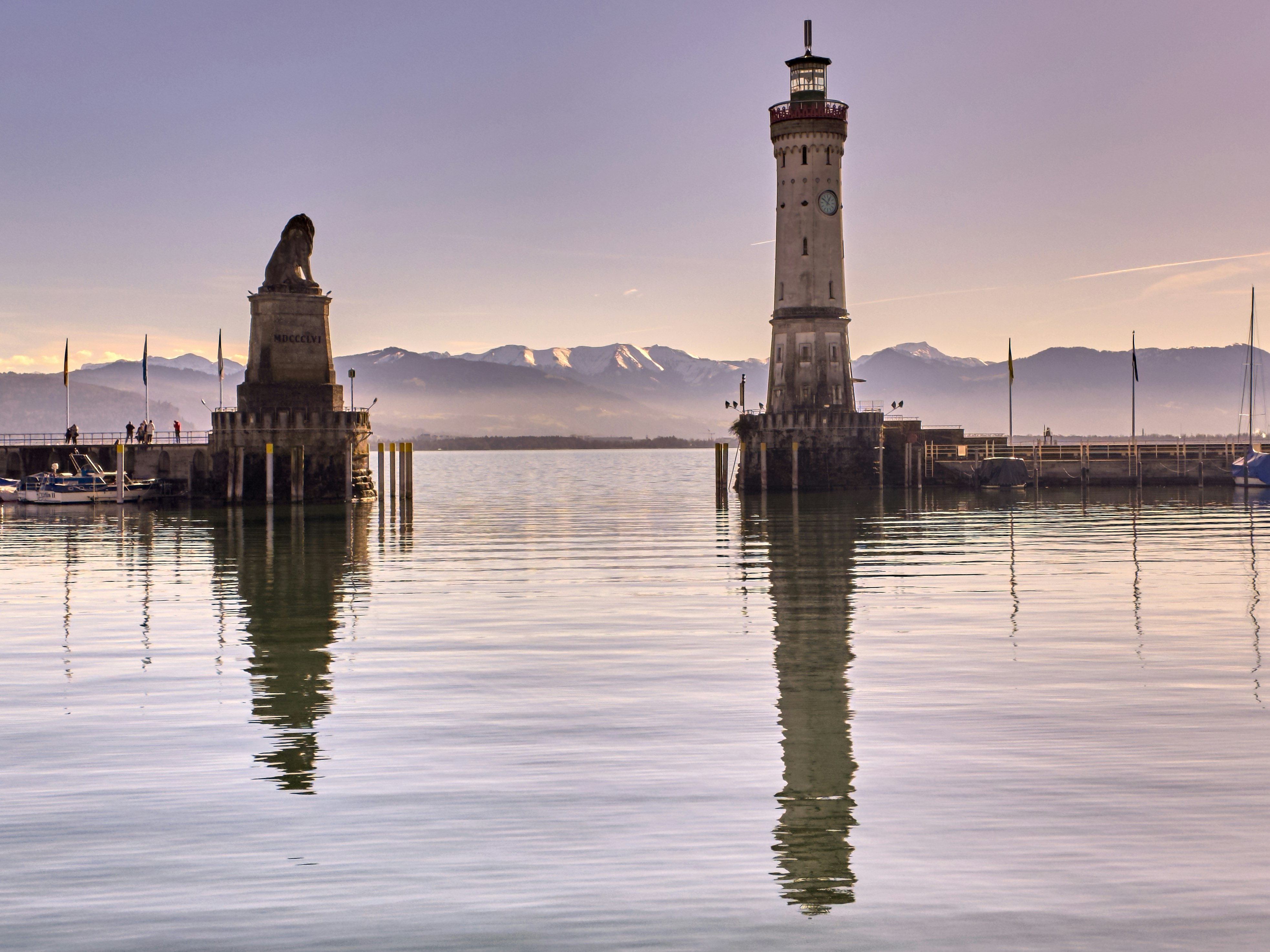 Lighthouse and lion statue reflecting in calm waters at sunrise, with distant mountains under a pastel sky.