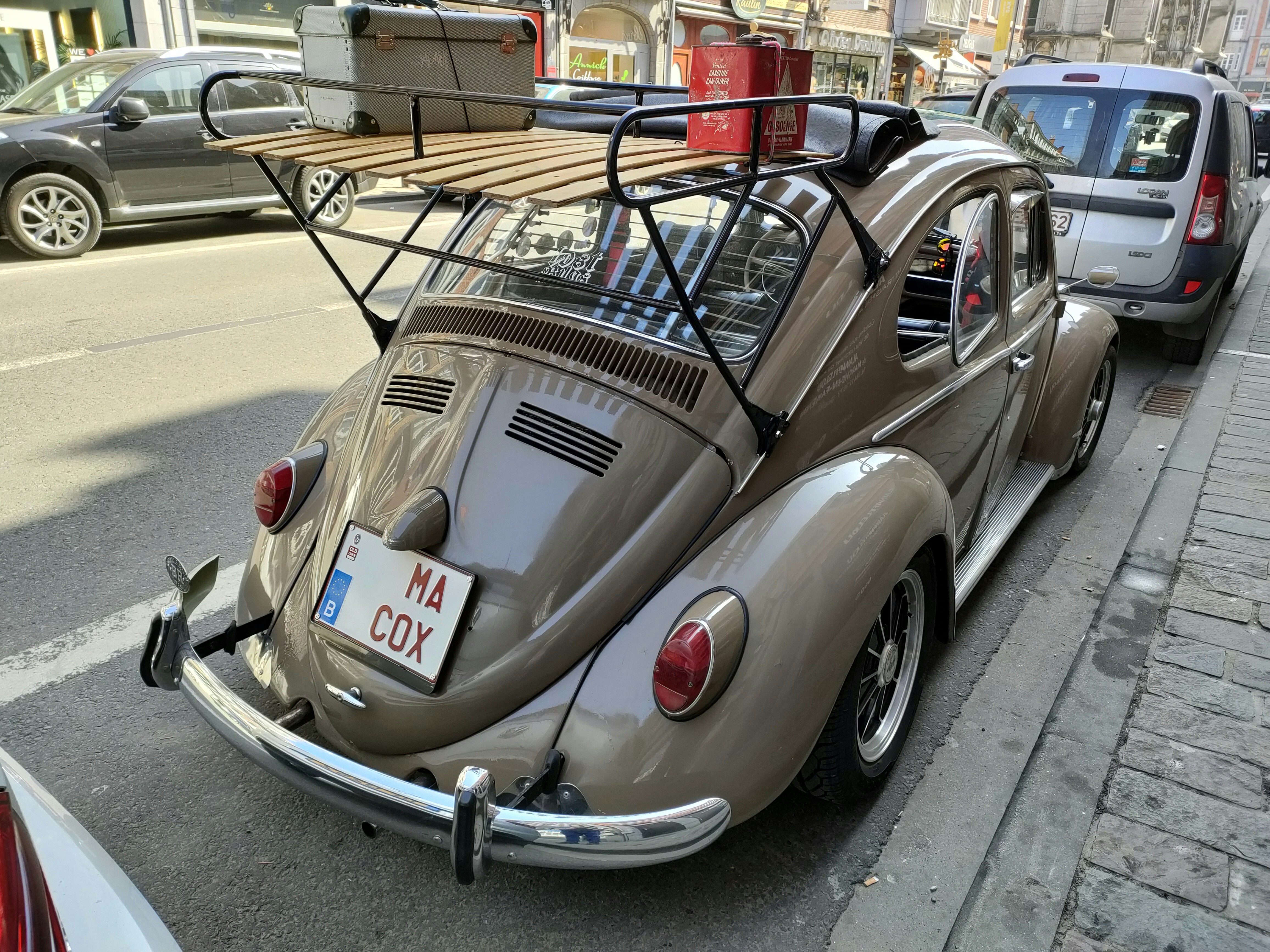 Beige vintage Volkswagen Beetle parked along a city curb with a metal rear luggage rack, captured in daylight street photography.