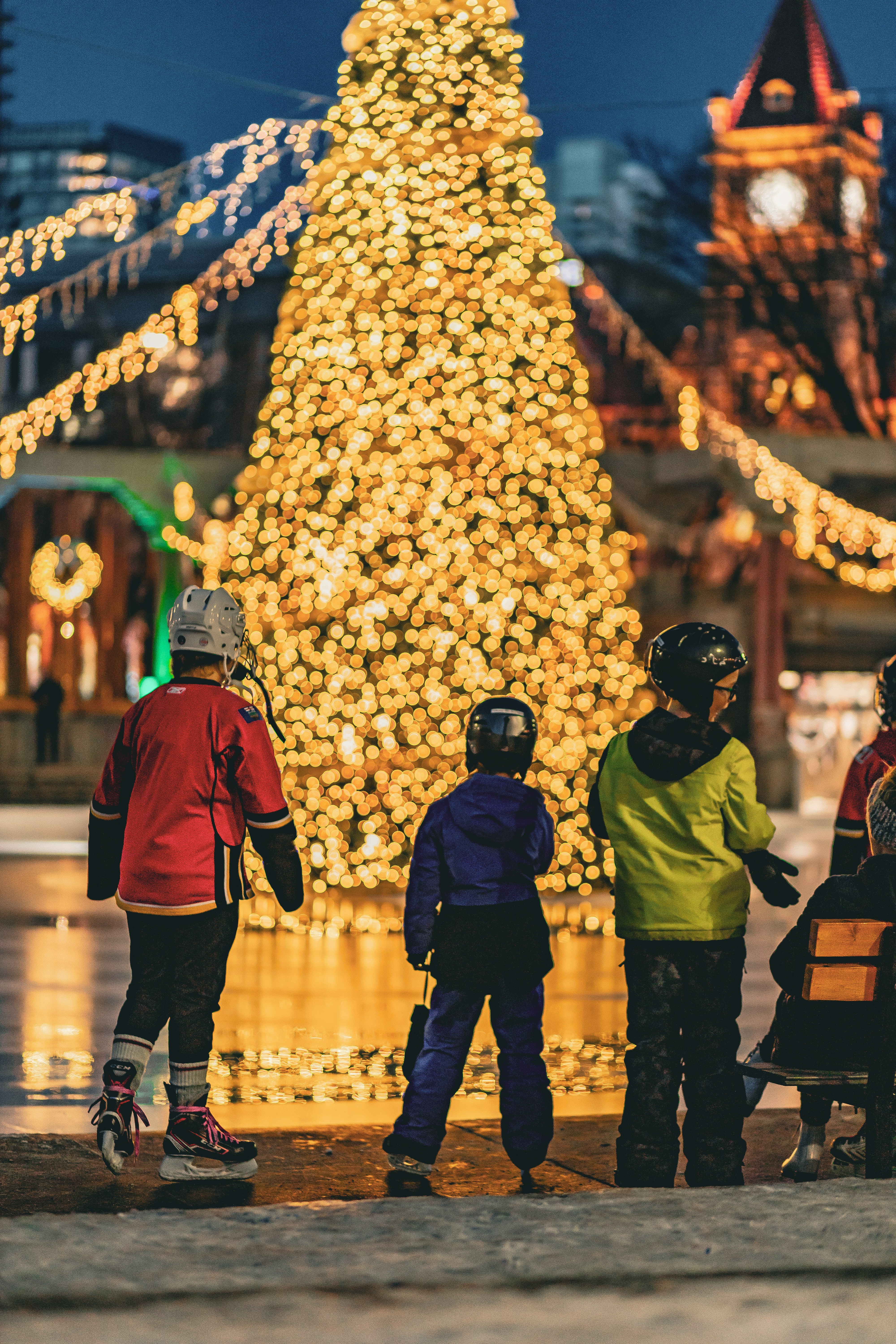 Children in colorful gear gaze toward a glowing Christmas tree in a festive plaza