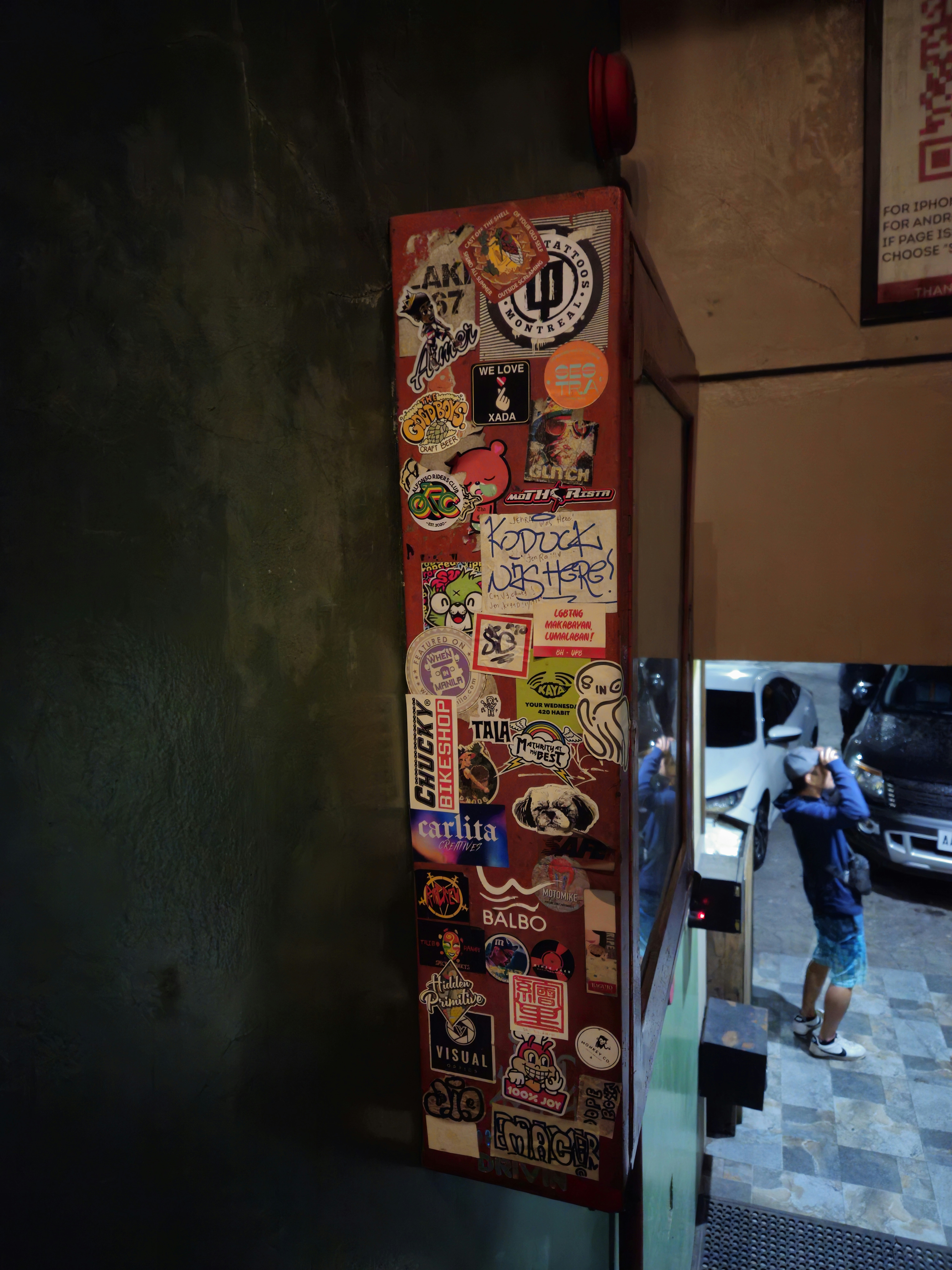 Dim stairwell with a vertical panel covered in colorful stickers beside a doorway; through the opening, a person stands near a parked car outside.