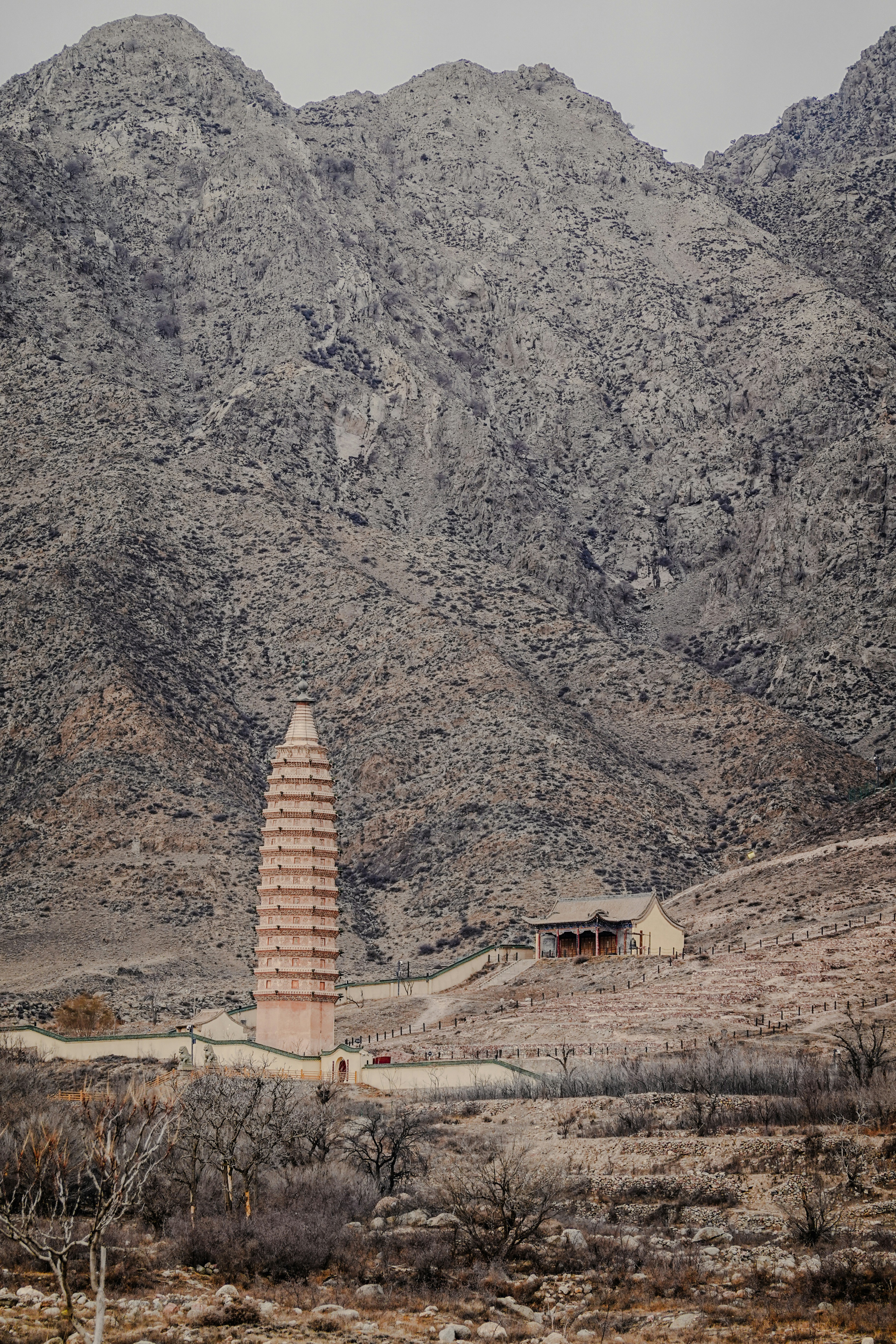 A tall pagoda stands prominently in a barren landscape, framed by rugged mountains under a cloudy sky. The scene conveys a sense of isolation and serenity.