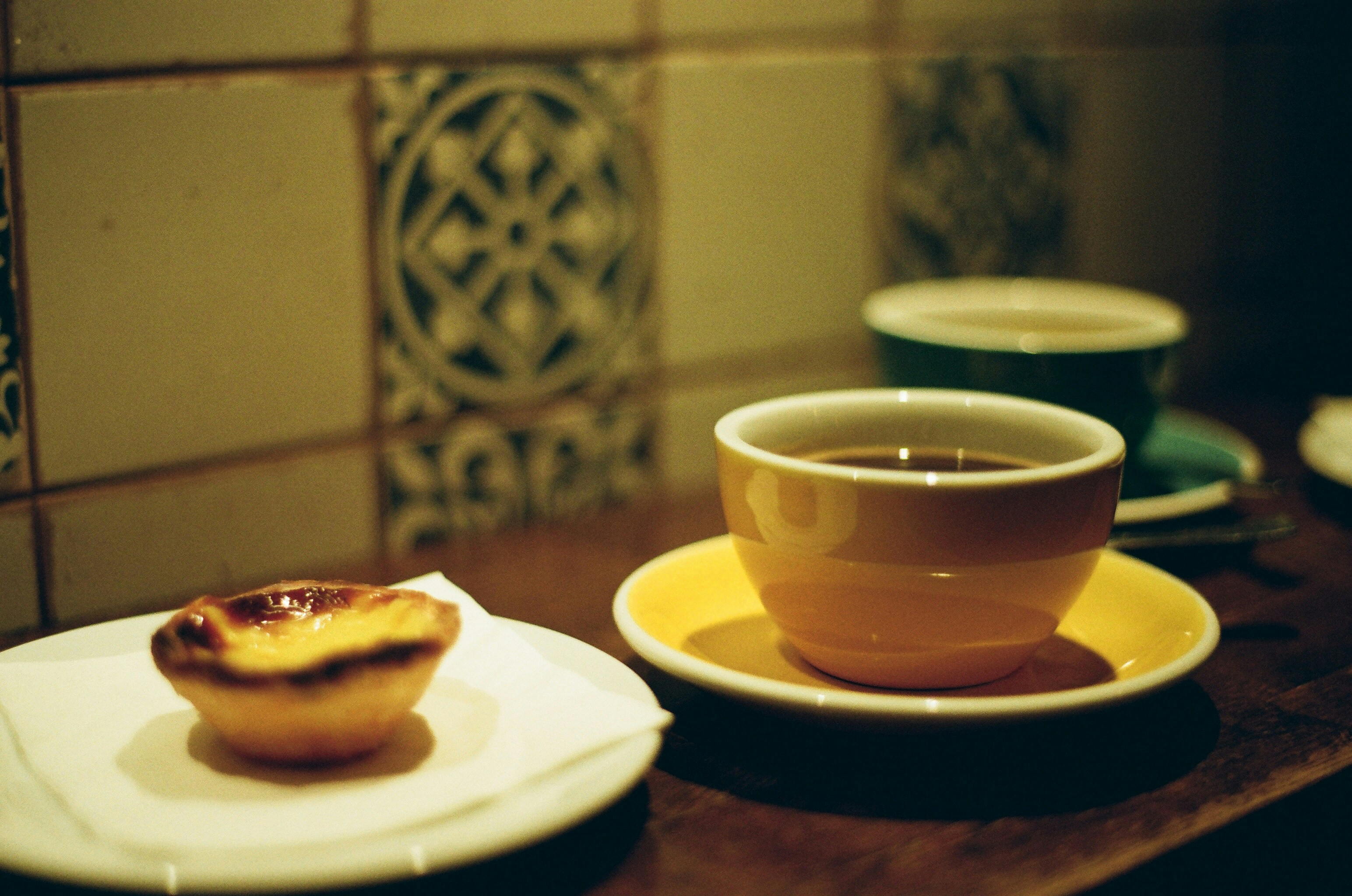 A warm pastel de nata on a napkin beside a steaming cup of coffee against a tiled wall.