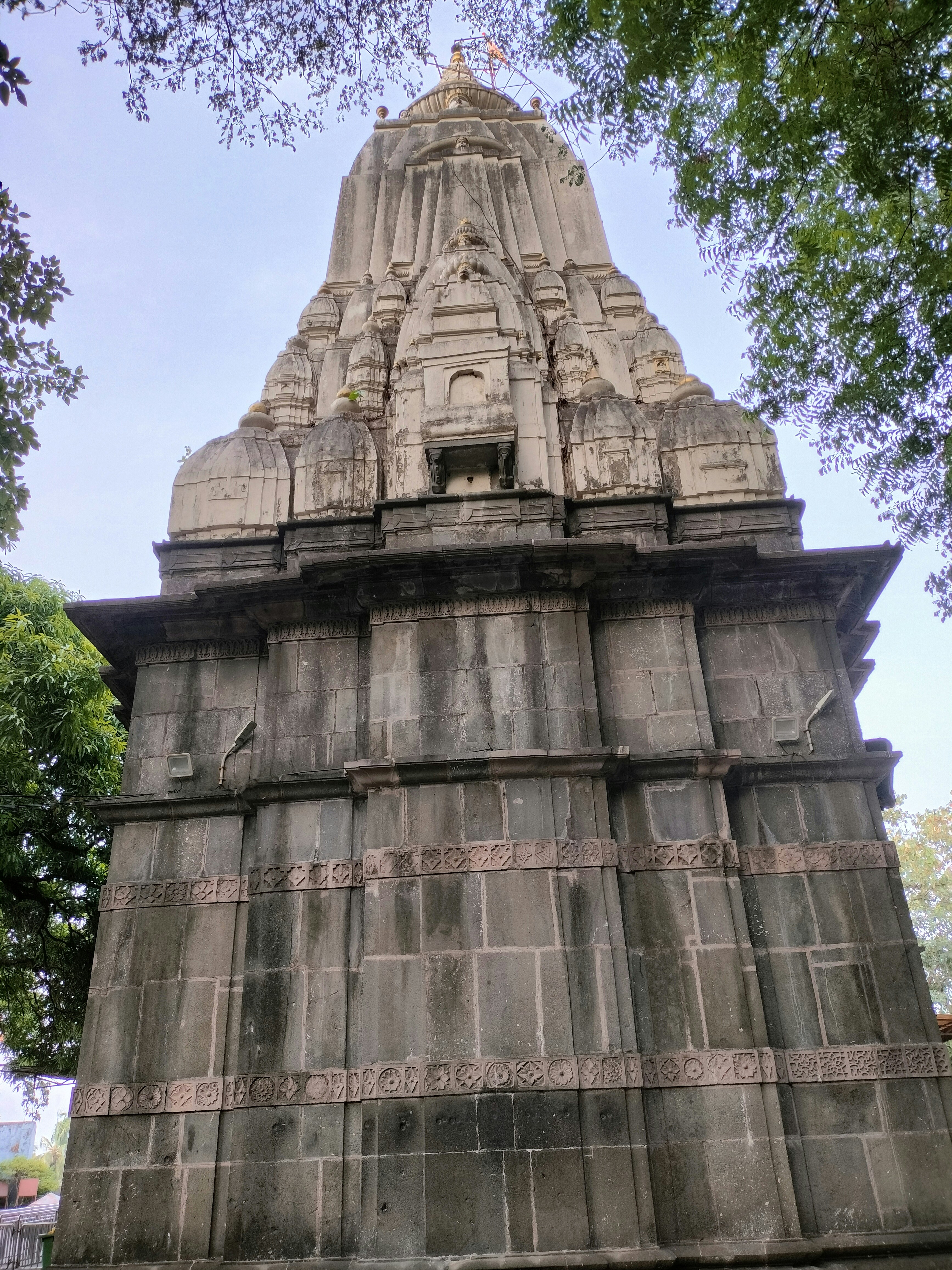 Weathered stone temple tower with layered, sculpted tiers rises under a leafy canopy. Photograph captures the masonry details and contrast with surrounding greenery.