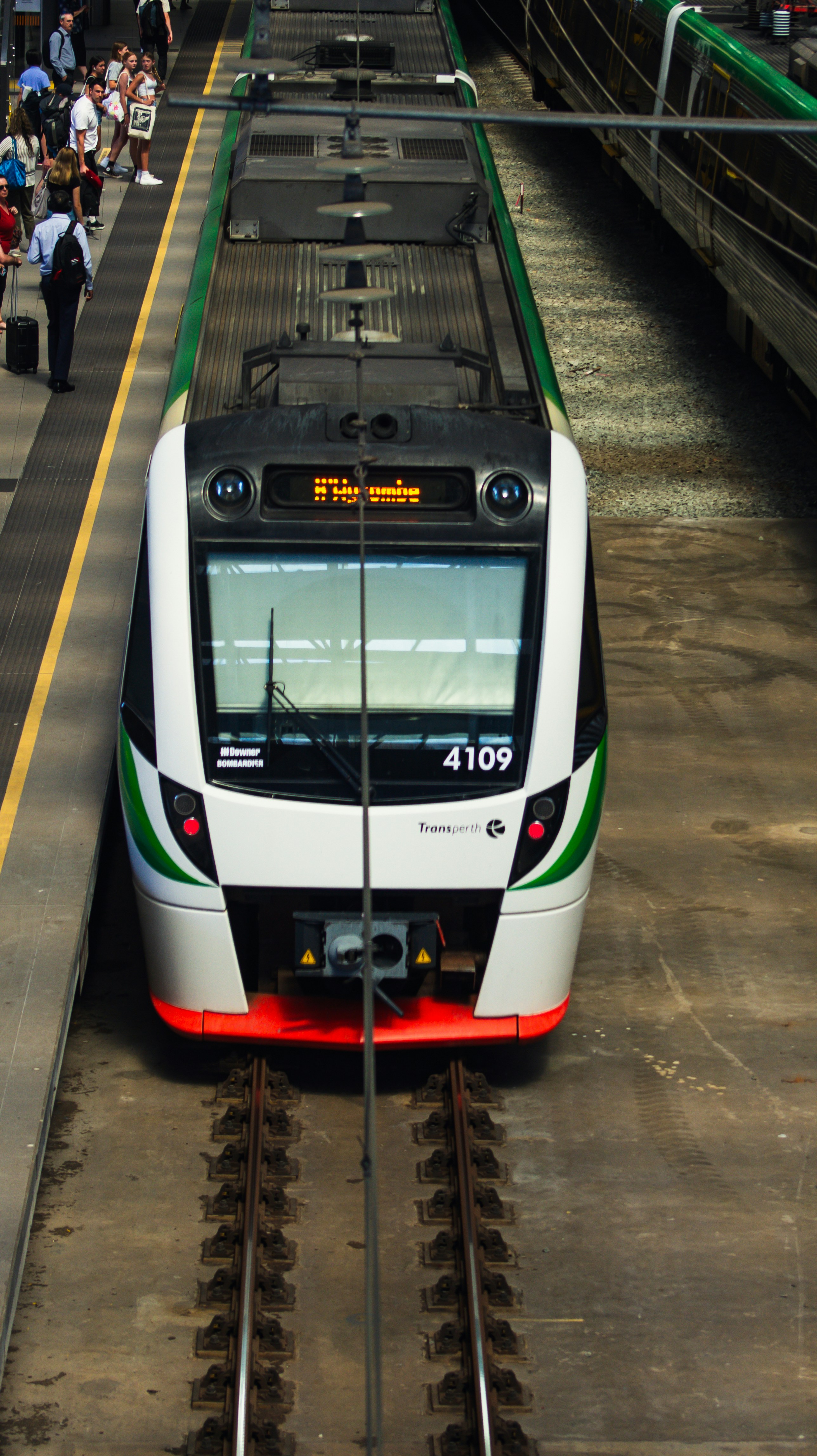 Front-on photograph of a modern electric train at a busy platform with passengers nearby. The sleek white-green-red nose dominates the frame, highlighting urban commuter rail branding.