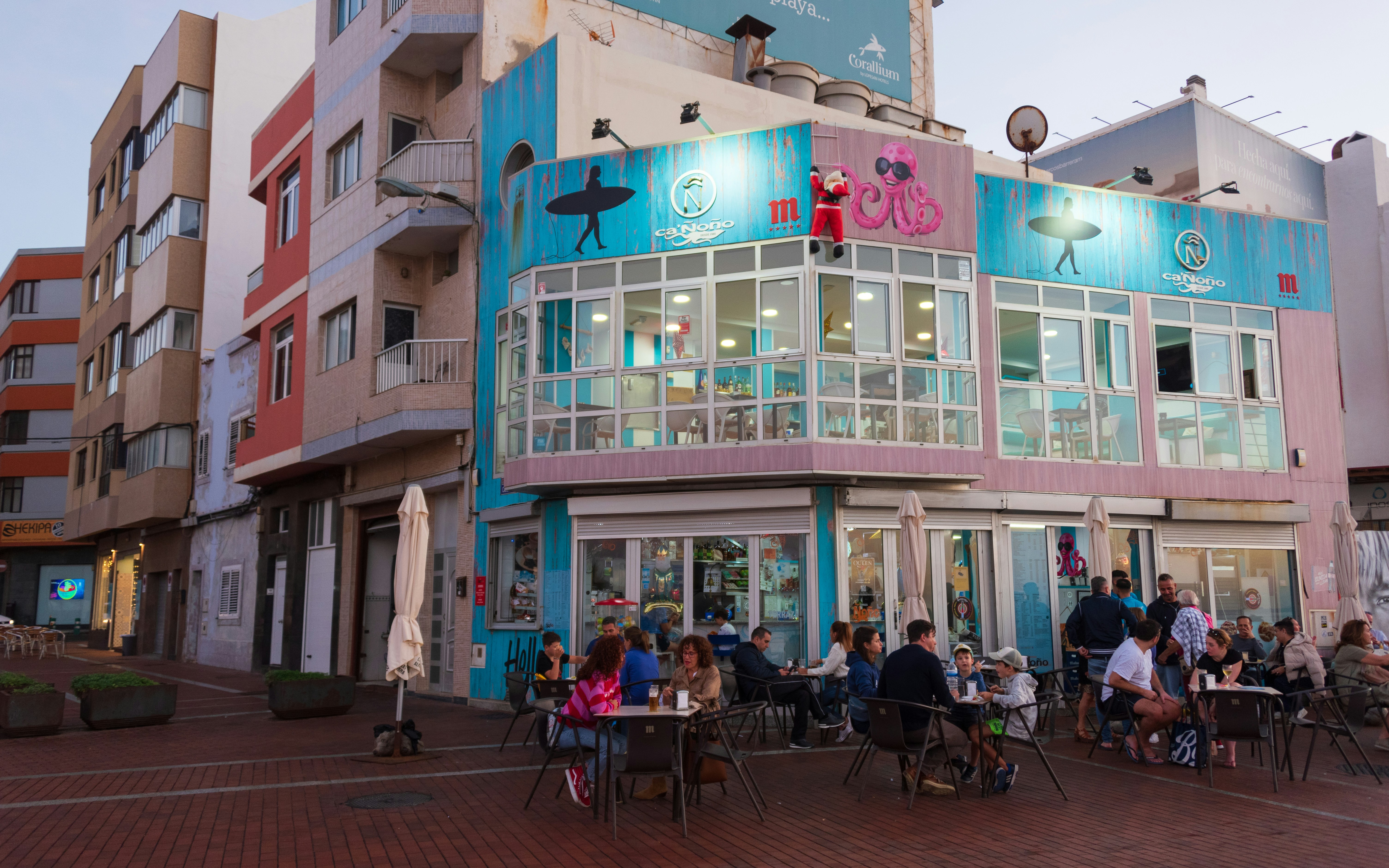 A lively bar in Las Palmas, Gran Canaria, with colorful murals featuring a surfboard and bicycle. Patrons enjoy drinks at outdoor tables on a red brick street, creating a vibrant and social atmosphere.
