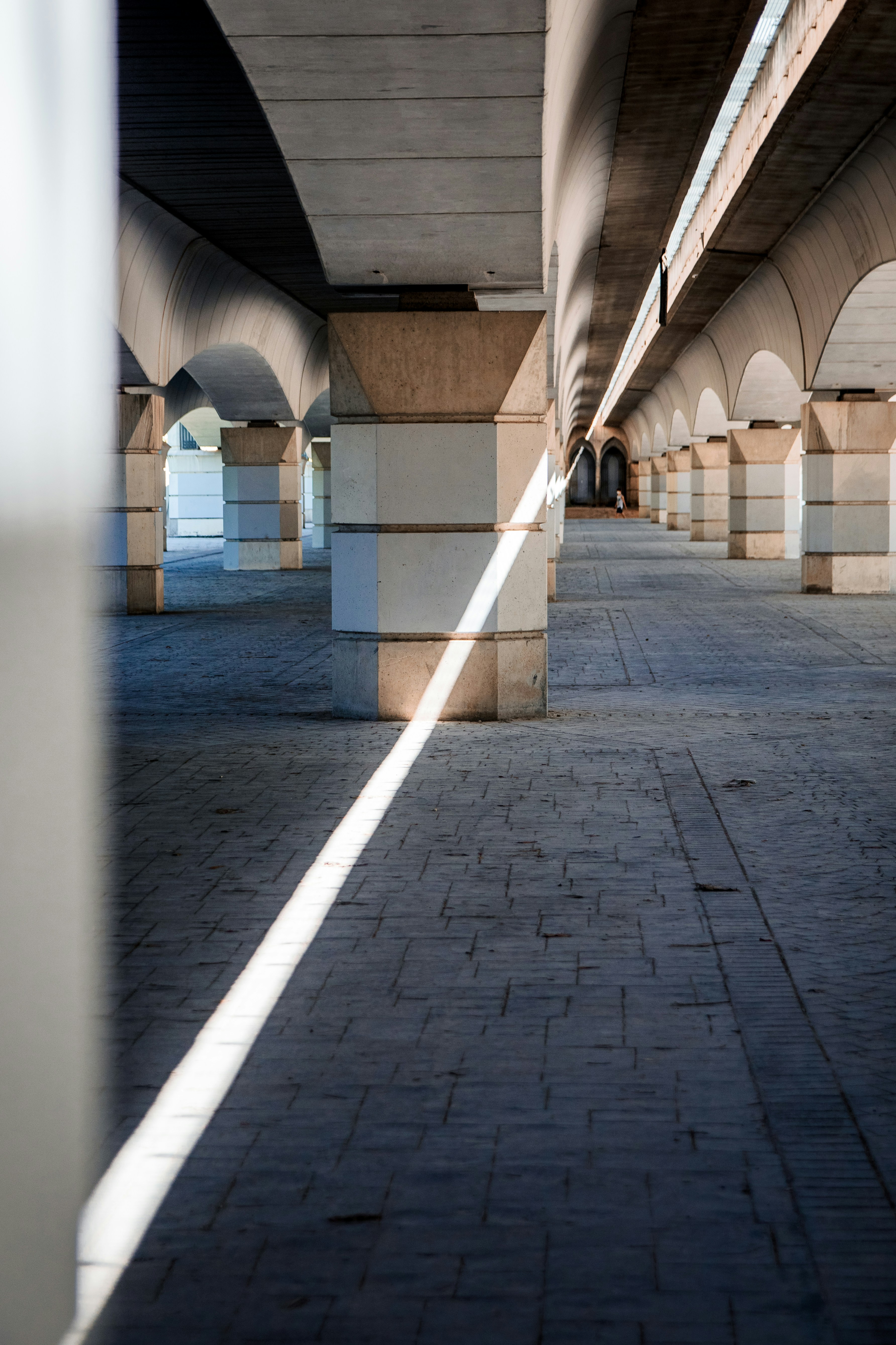 Minimalist architectural scene beneath a large concrete bridge or viaduct, featuring repeating arches, strong perspective lines, and a dramatic diagonal beam of sunlight across the pavement and support column. The quiet, empty walkway creates a modern, geometric, urban atmosphere—ideal for concepts like structure, infrastructure, symmetry, and solitude.