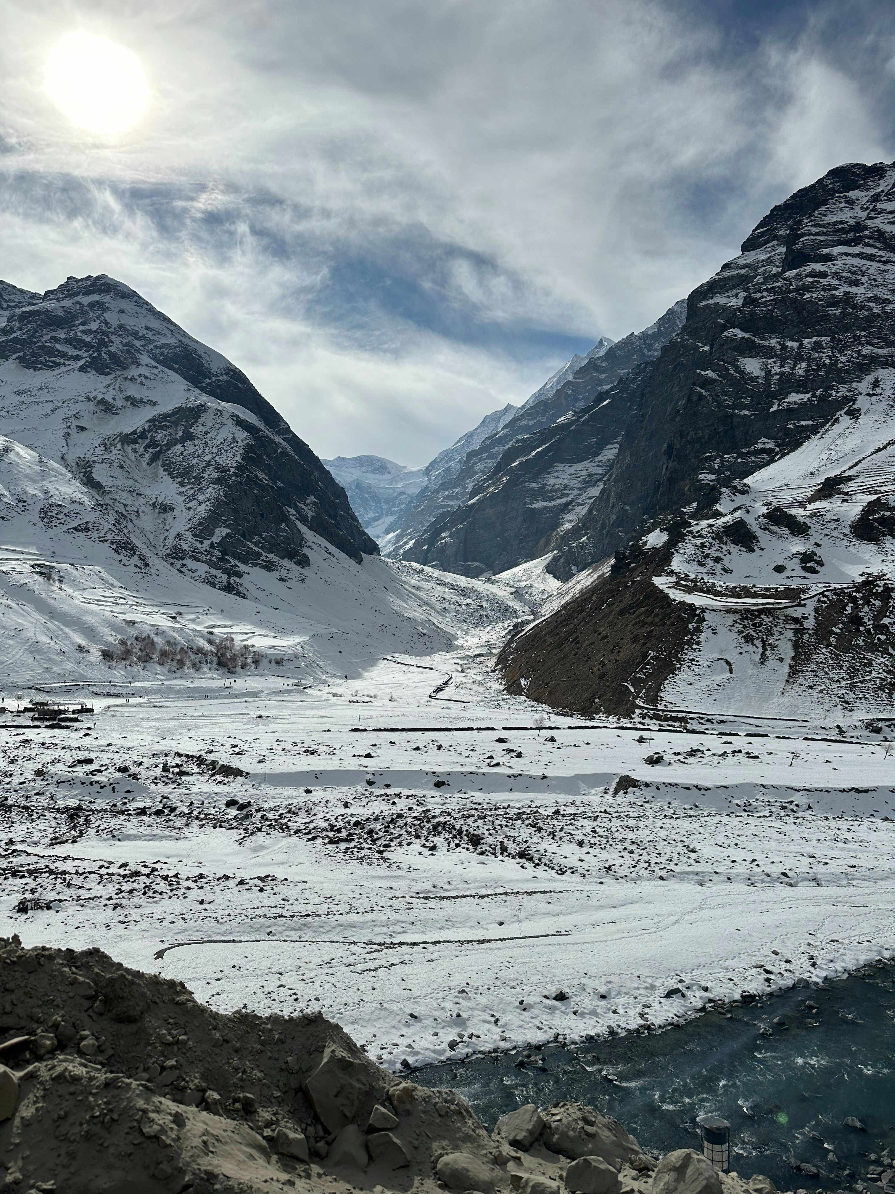 Snow-covered valley flanked by towering mountains under a bright sun, with a winding river cutting through the landscape.