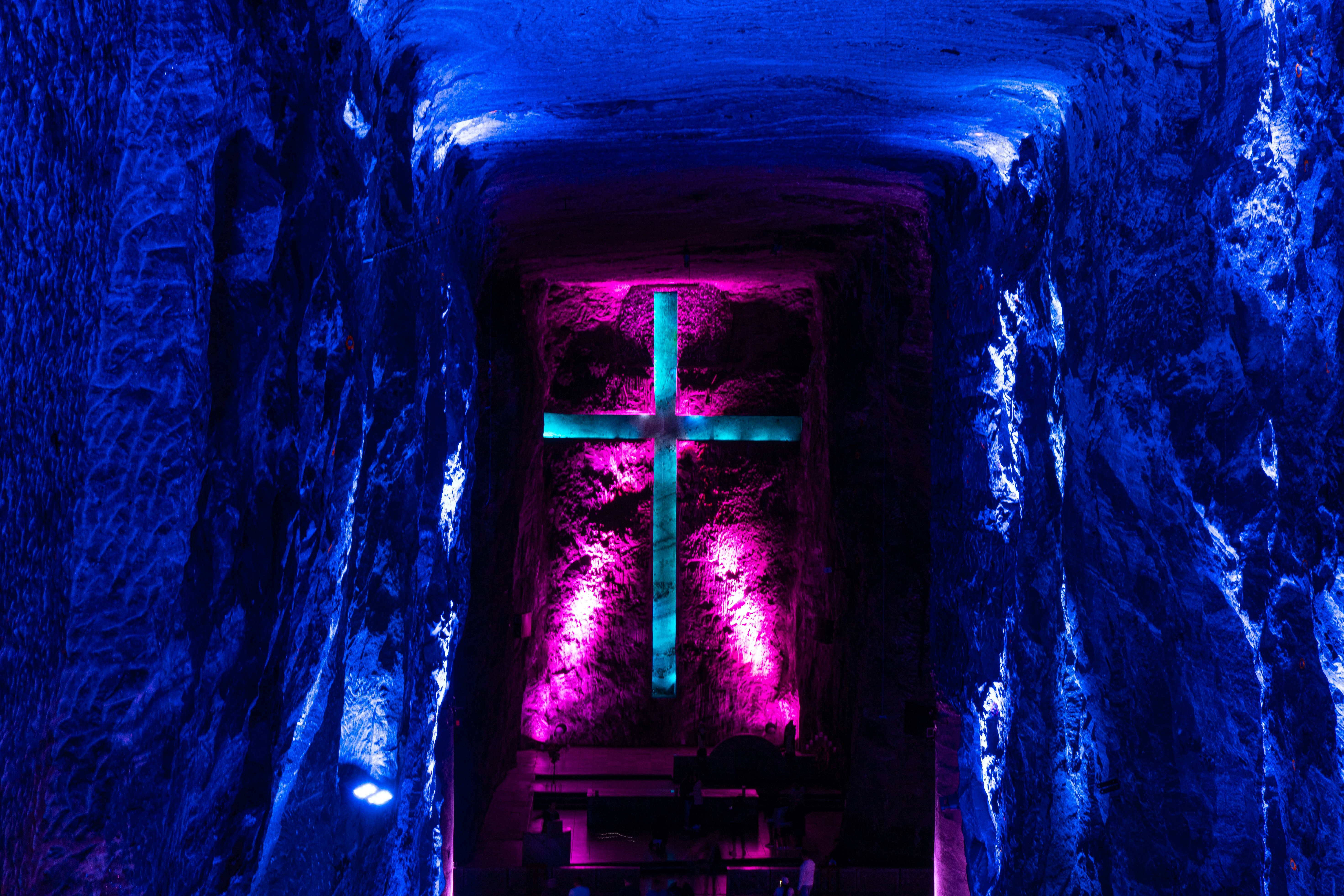 Vibrant neon cross glowing in a dimly lit cavern with blue and purple hues.