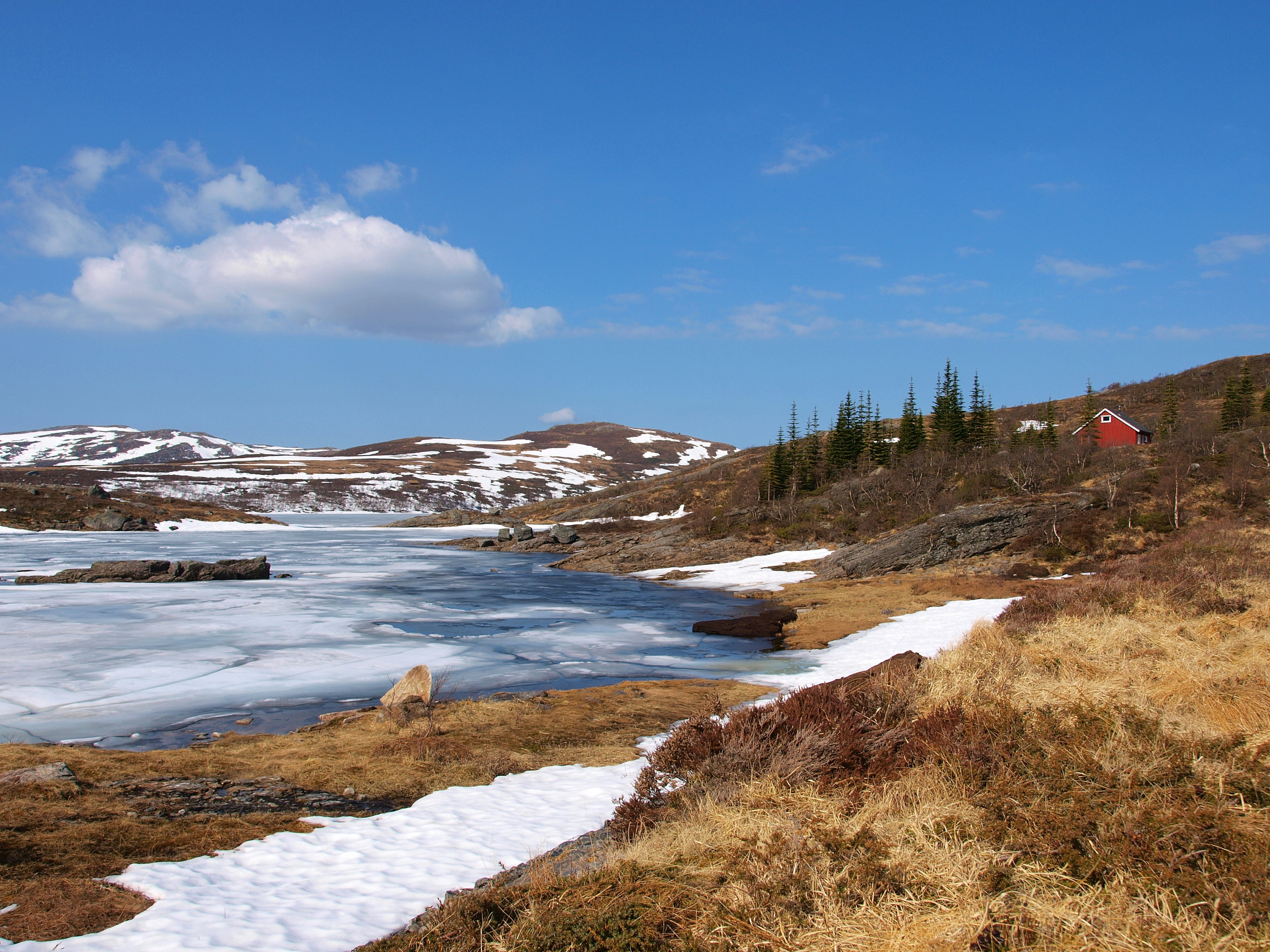 Snow-dusted hills cradle a partially frozen lake with brown grasses in the foreground and a red cabin perched on the hillside under a clear blue sky.