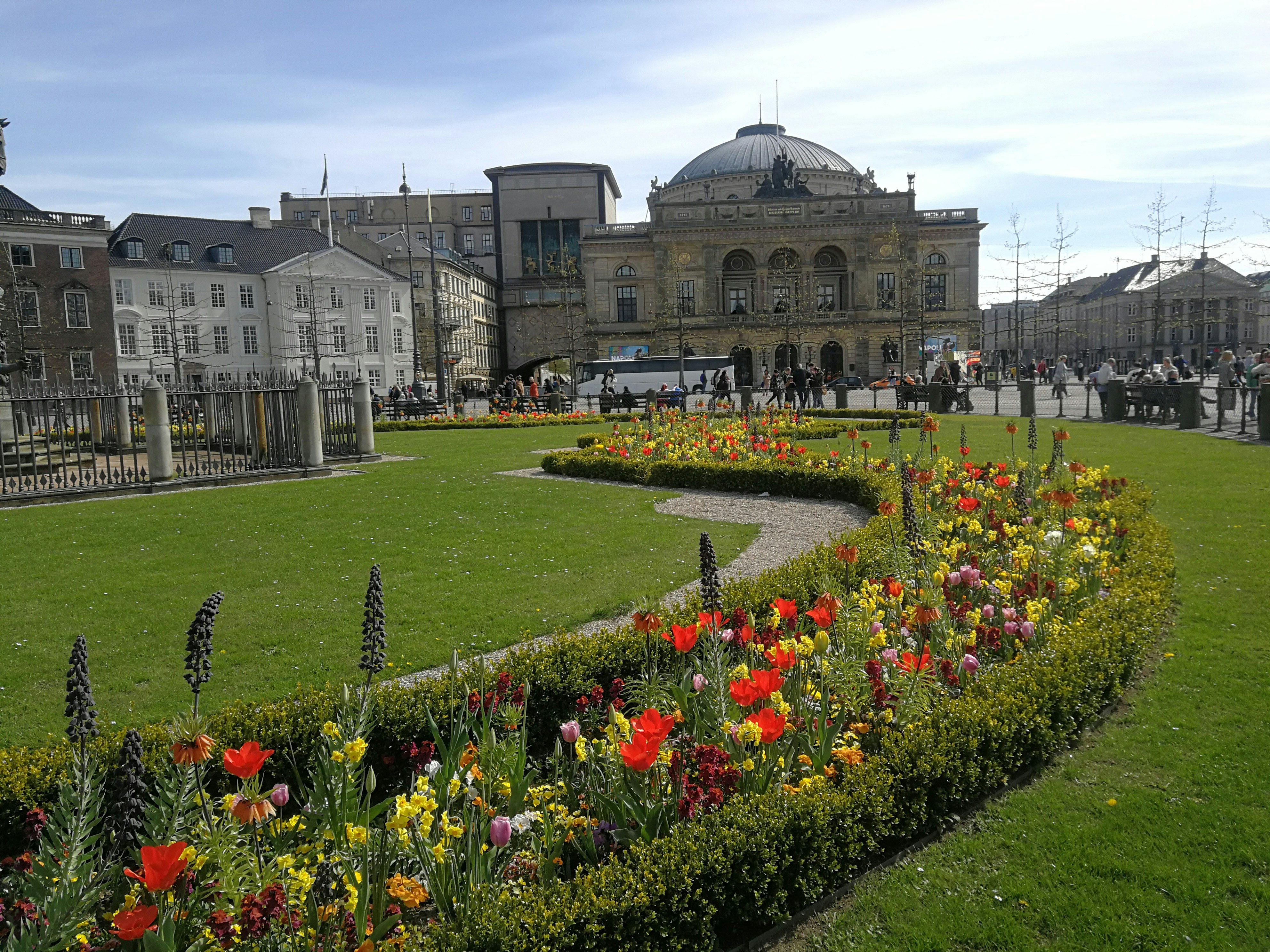 Vibrant flower beds in a city square with an ornate building in the background, bustling with visitors. 