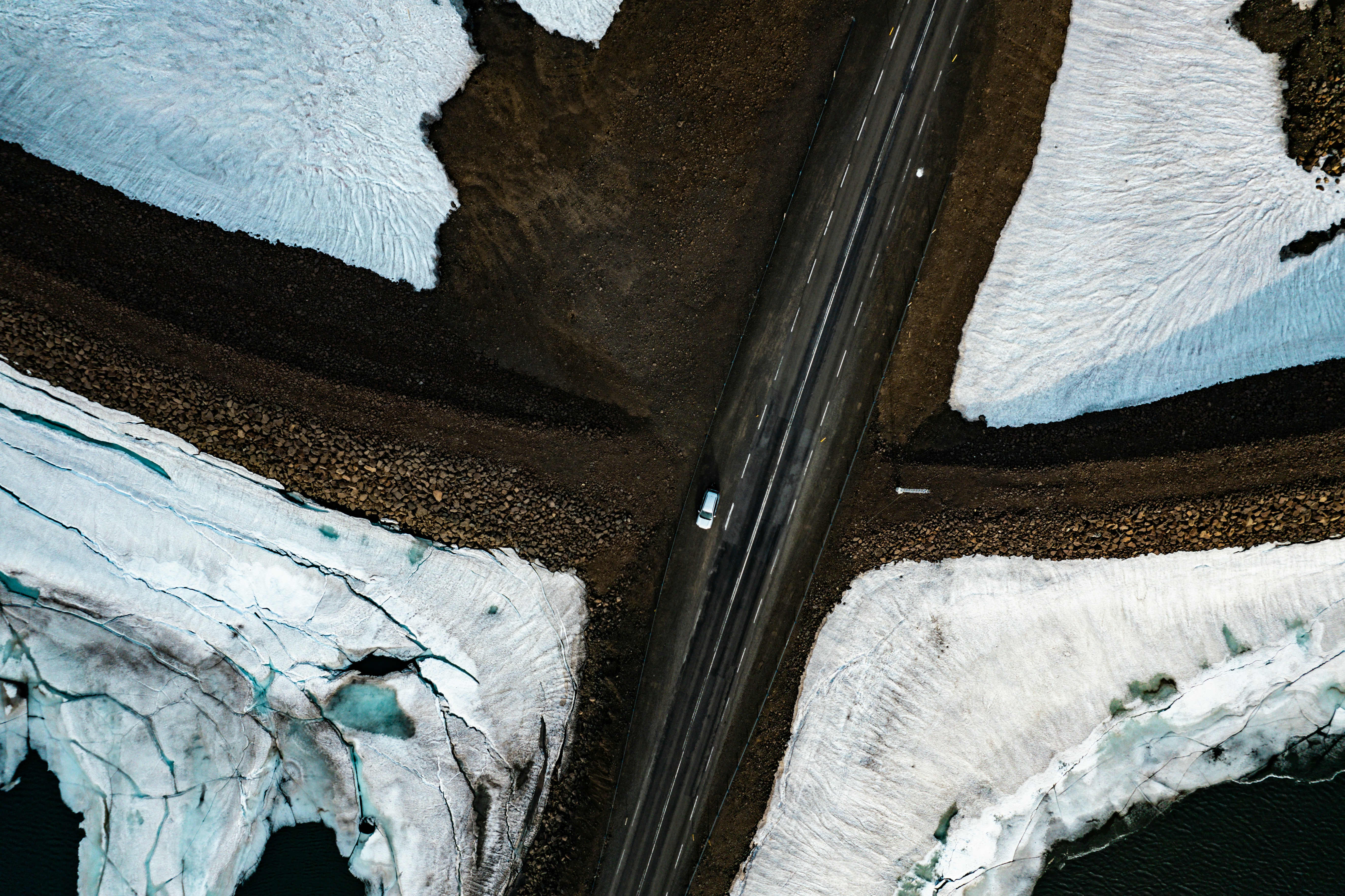 A bird's-eye view captures a solitary vehicle traversing a road that slices through a landscape of snow, ice, and rocky terrain, illustrating a stark contrast between human endeavor and the raw power of nature.