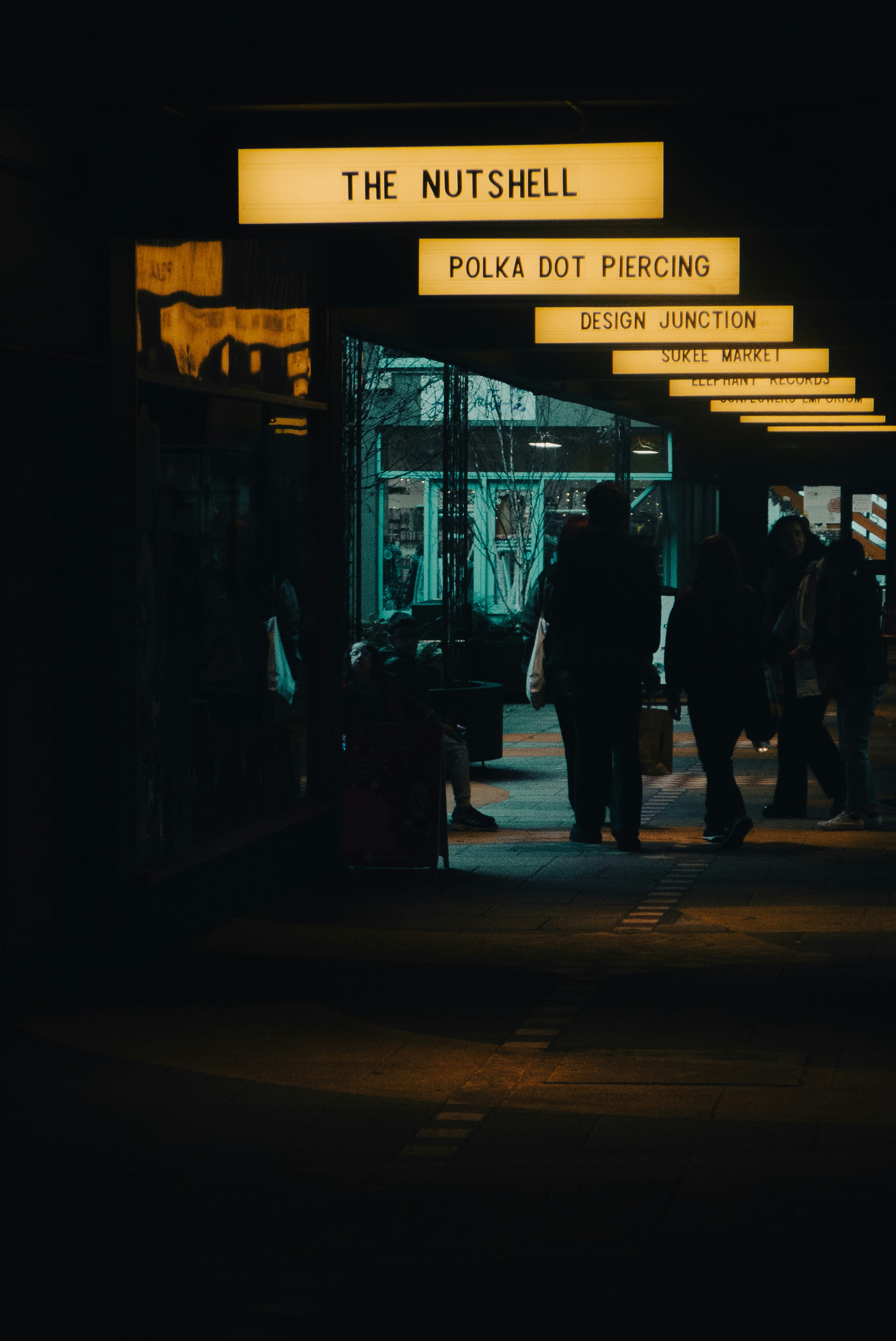 Dimly lit urban alley with silhouetted figures and glowing storefront signs.