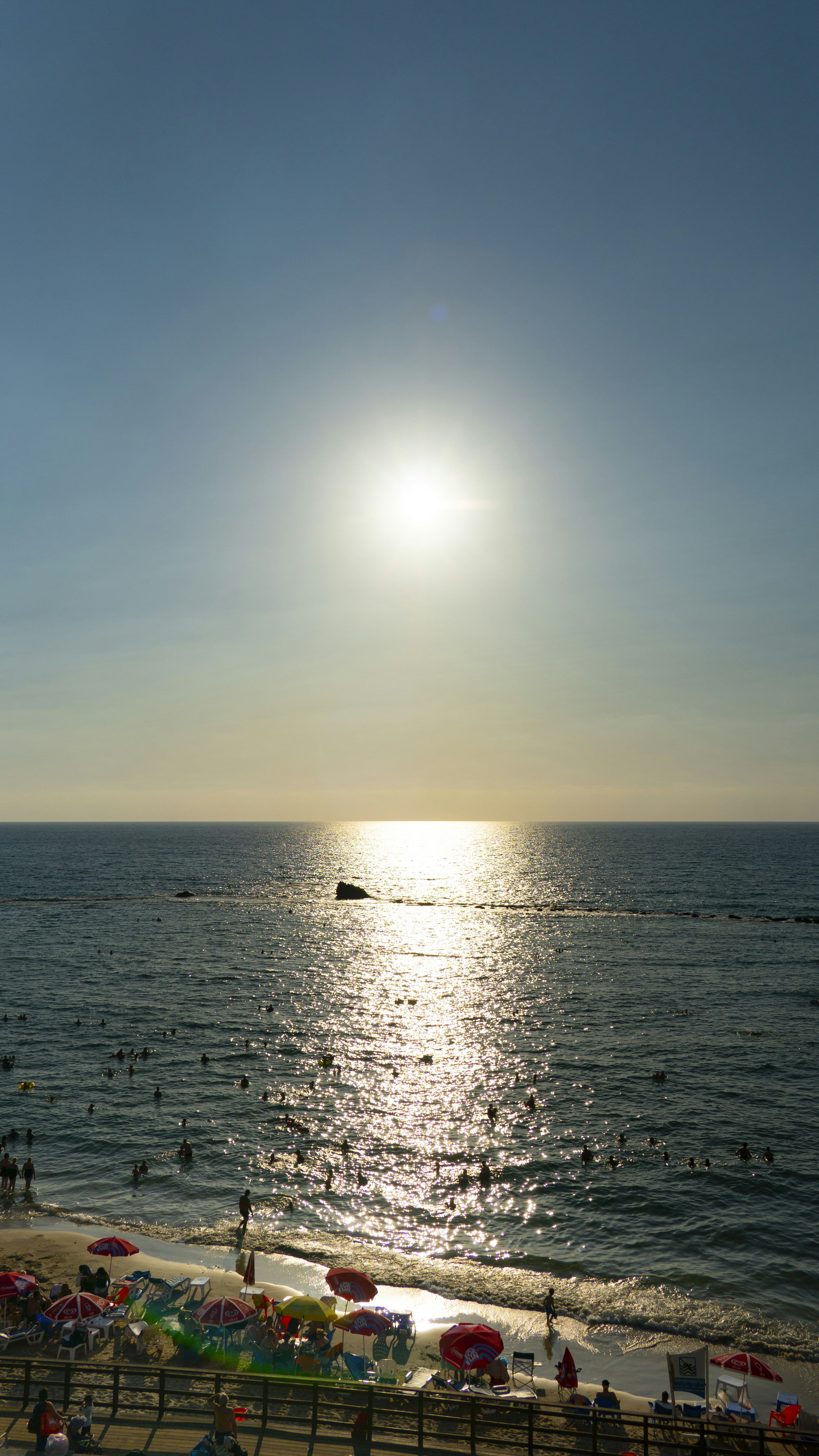 Beachgoers under colorful umbrellas along Bat Yam's sun-drenched coastline, with the sun reflecting on the ocean.
