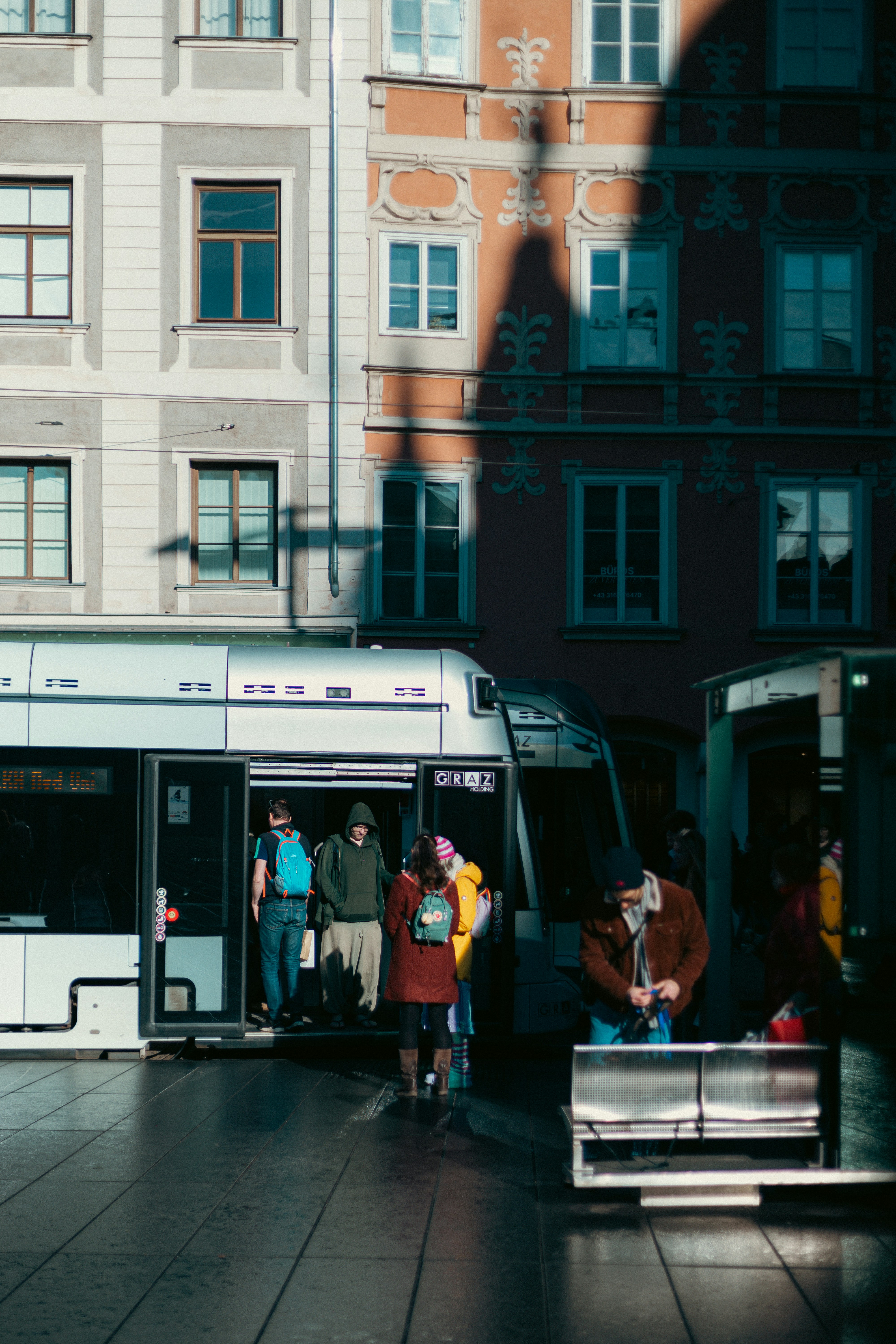 Shadow Crossing the Facade: Commuters at a Sunlit Tram Stop