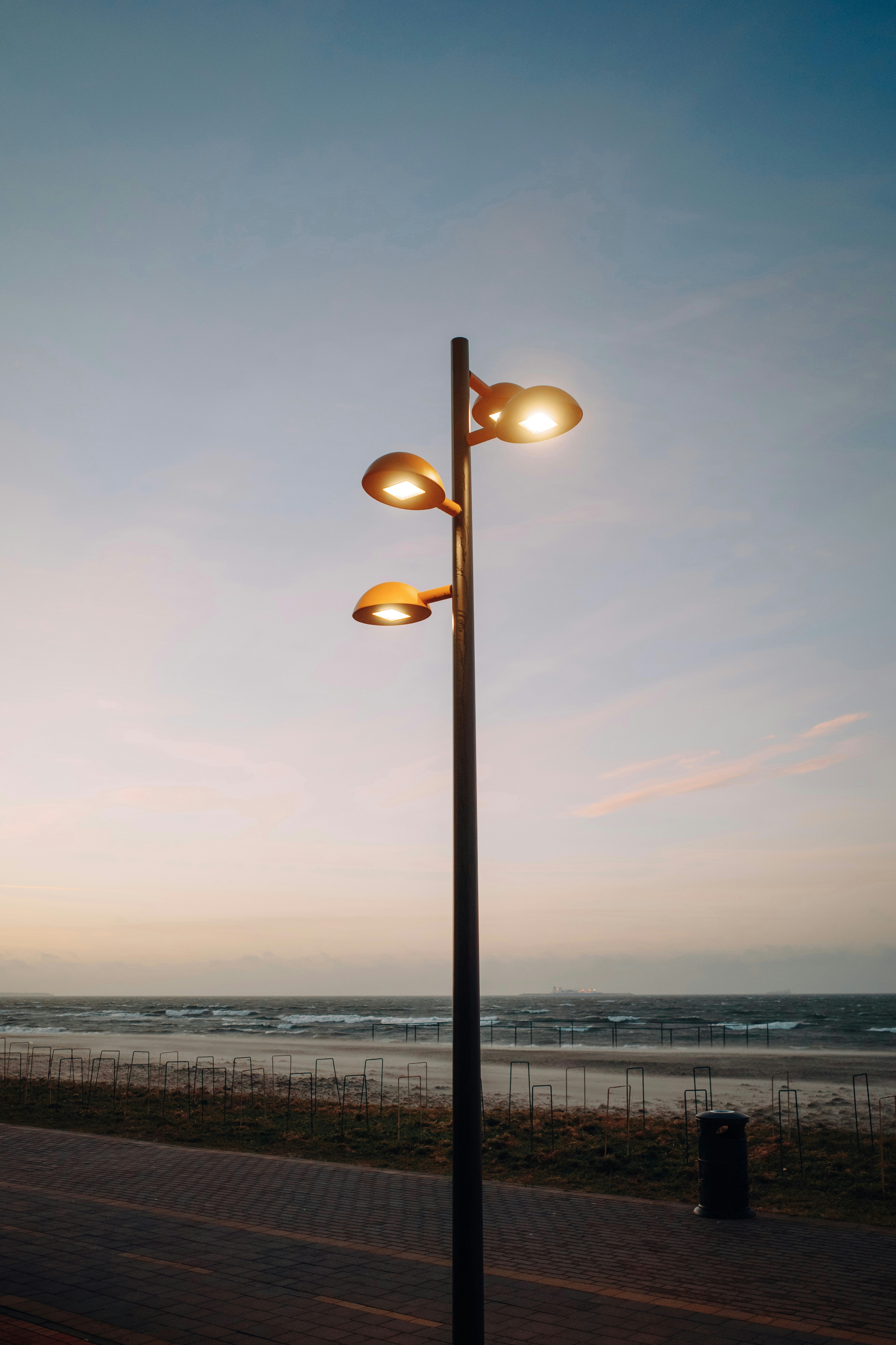  twilight beacon: three lamps on a lone boardwalk lamp against a sea‑front sky