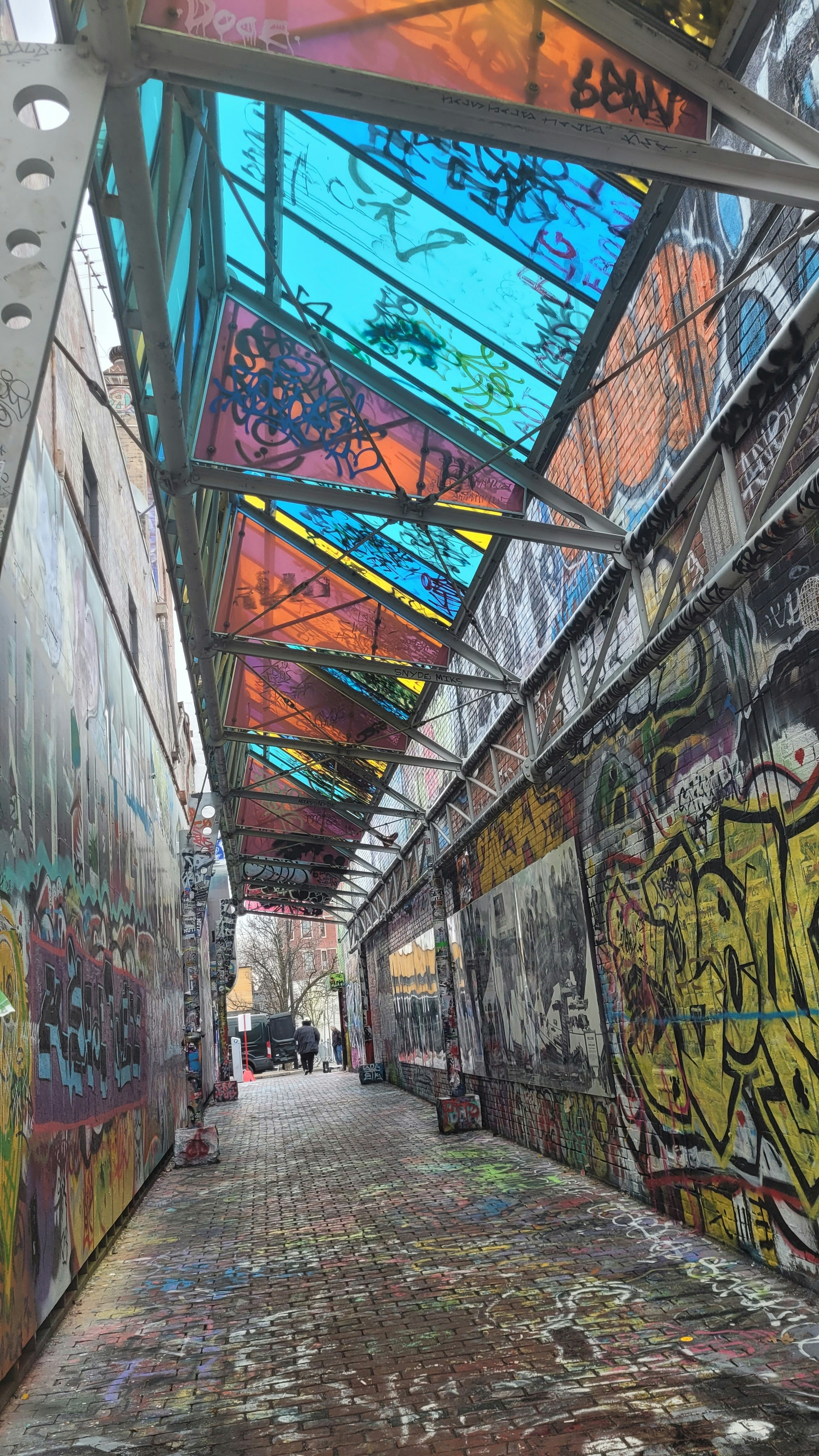 Colorful graffiti-lined alleyway beneath a striking glass canopy, showcasing a blend of urban art and architectural design.