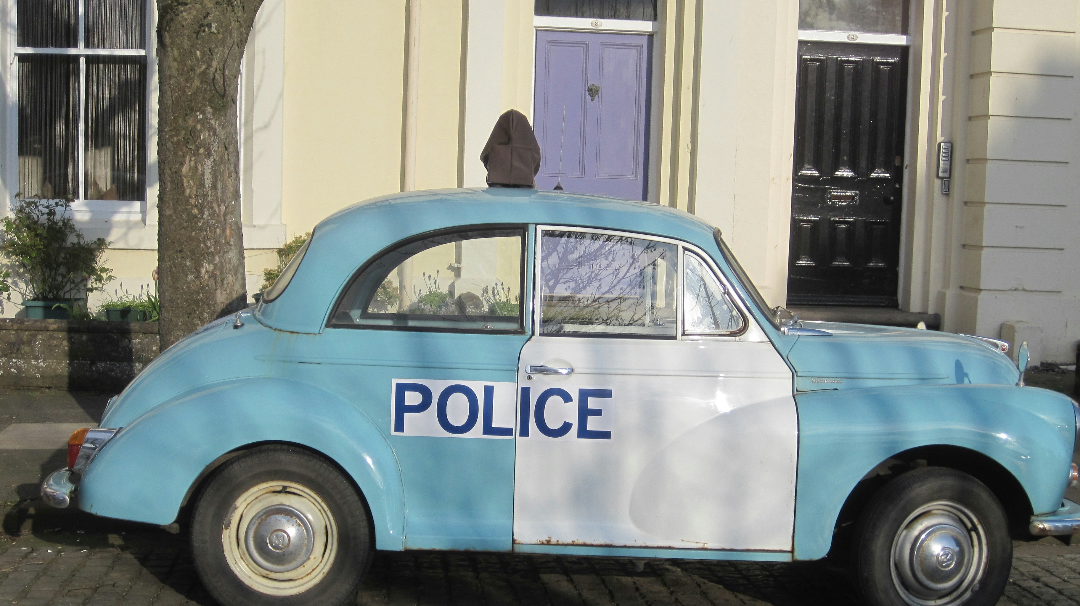 A vintage light-blue police car with a white central door panel sits parked on a curb in front of a pale yellow building. The retro vehicle anchors a calm street scene.
