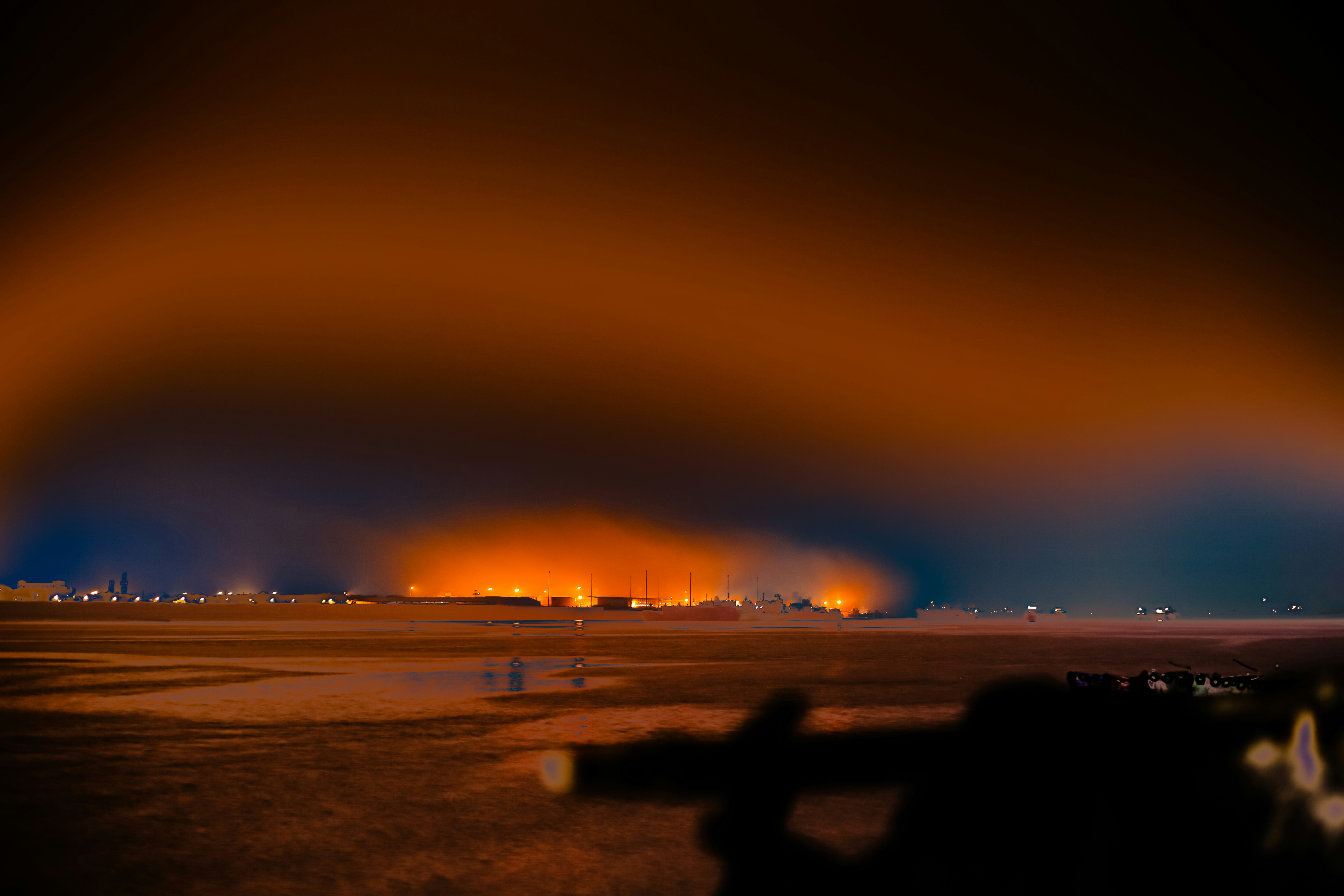 Orange and black sky reflecting on a calm body of water with distant industrial lights.