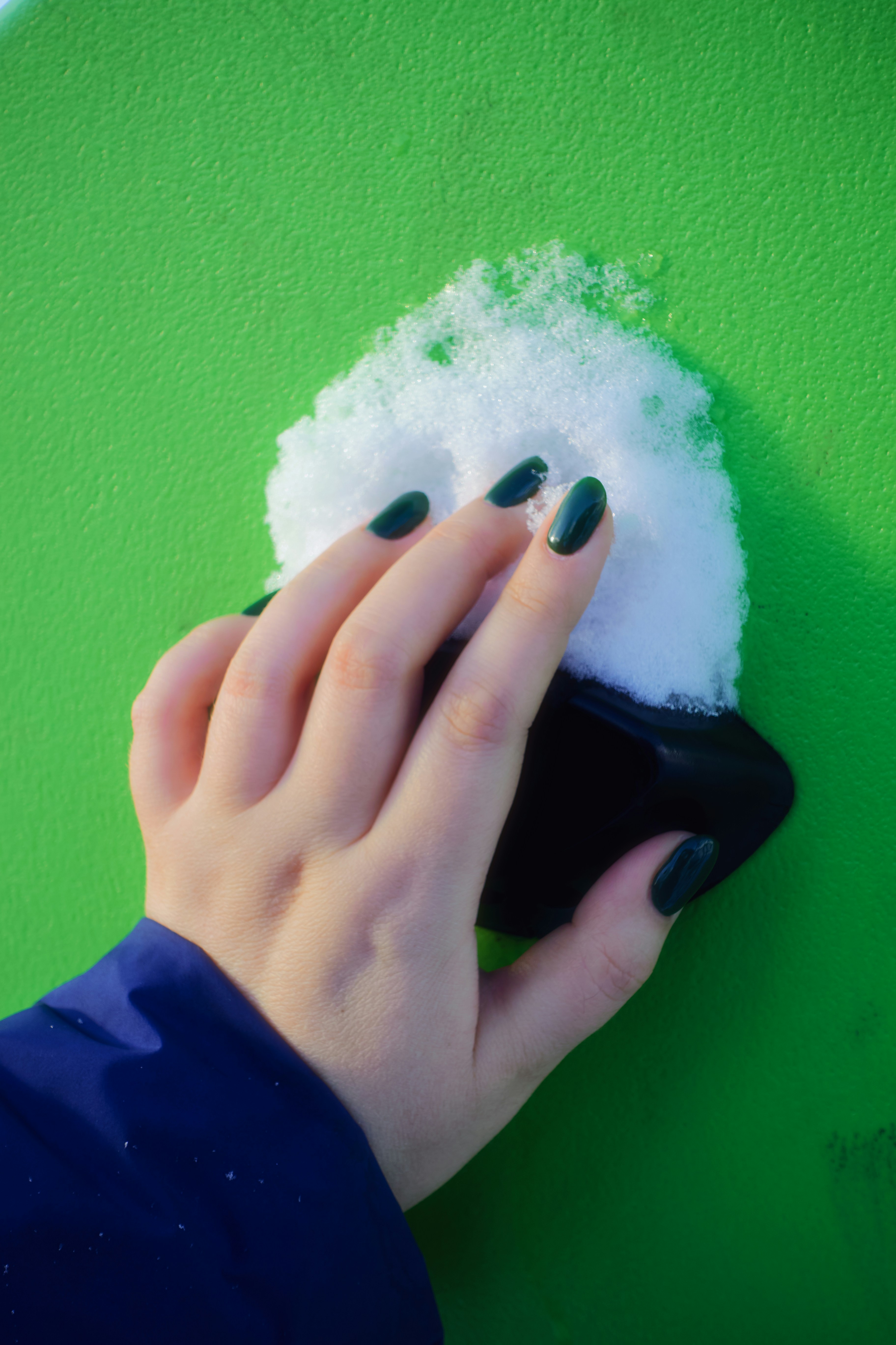 Close-up of a pale hand with dark teal nails gripping a black handle dusted with snow against a vivid green surface.