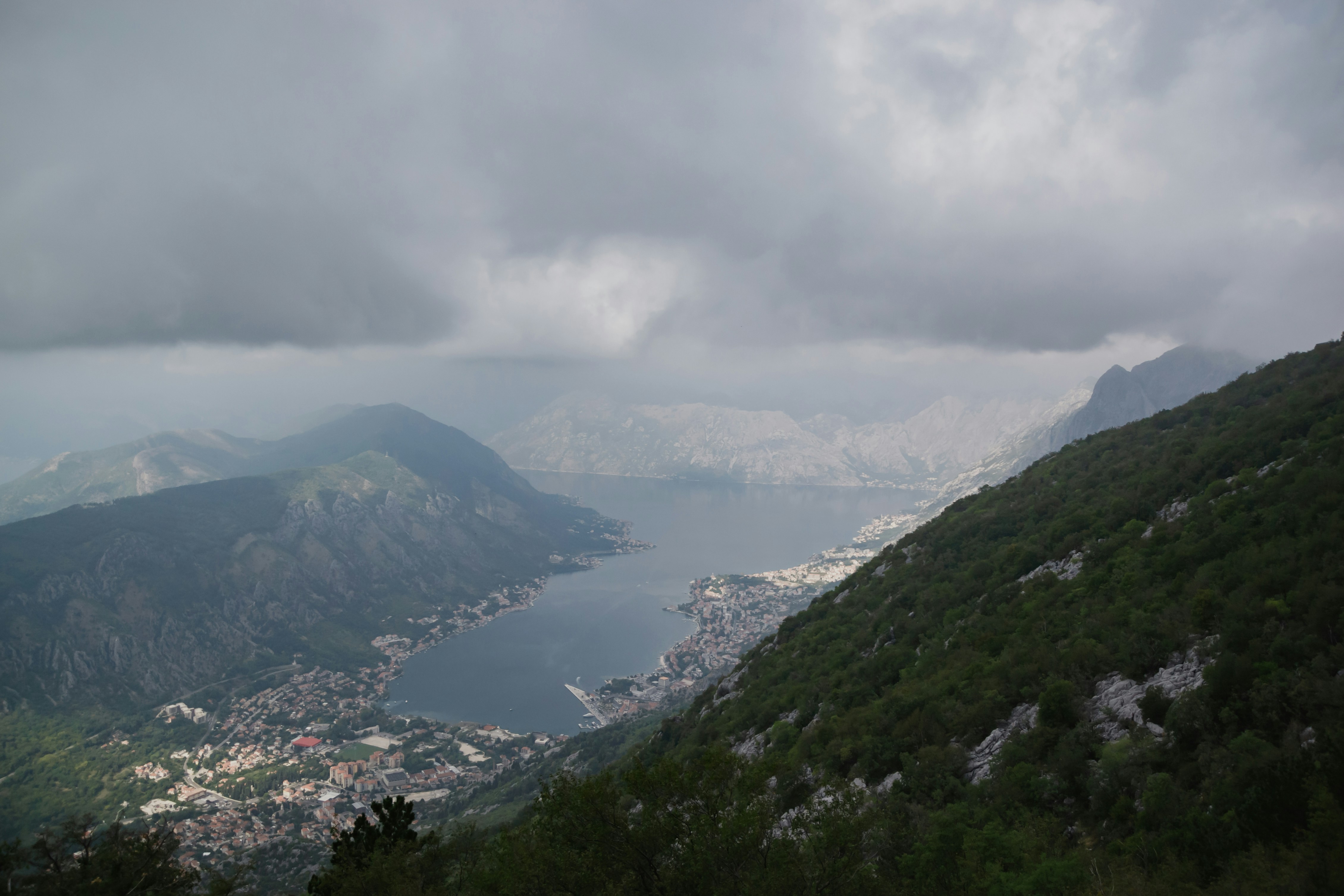 A panoramic view of a coastal town nestled between mountains under a moody sky. The interplay of light and shadow enhances the natural beauty of the landscape.
