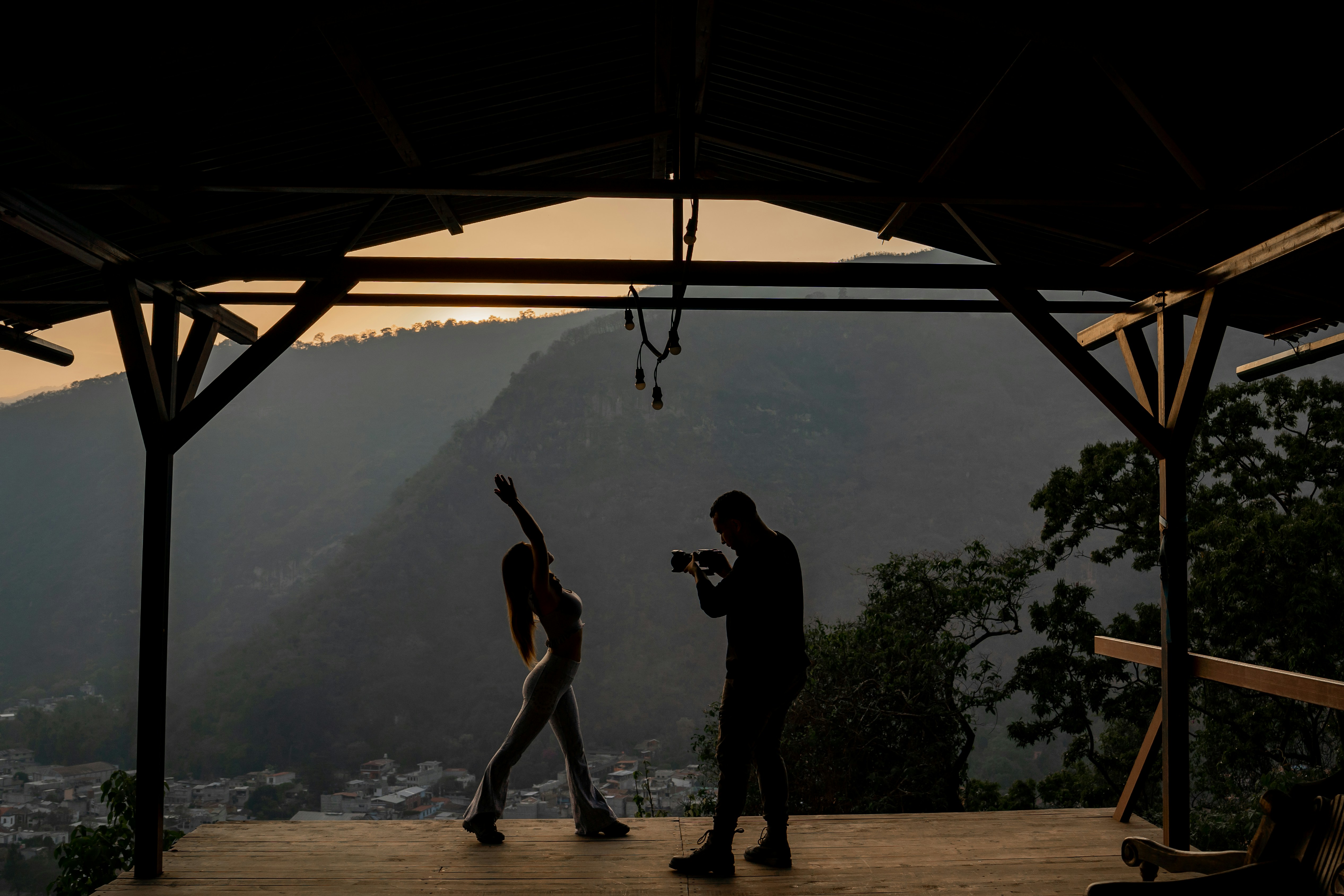 Dancer striking a pose in silhouette against a mountainous backdrop during sunset, while a photographer captures the moment.