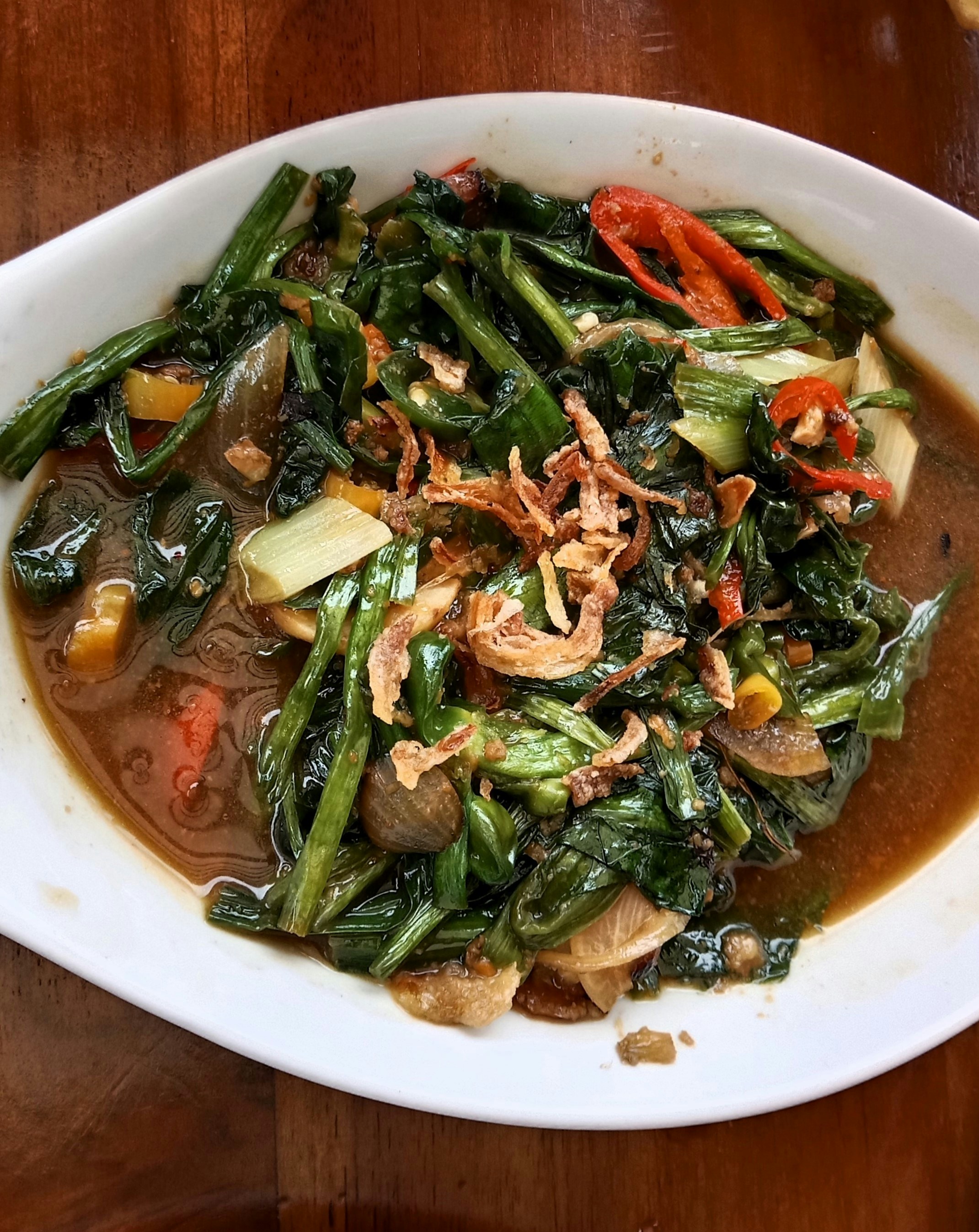 Vibrant stir-fried greens with colorful peppers and crispy shallots, served in a white dish. A savory sauce adds depth to this flavorful dish.