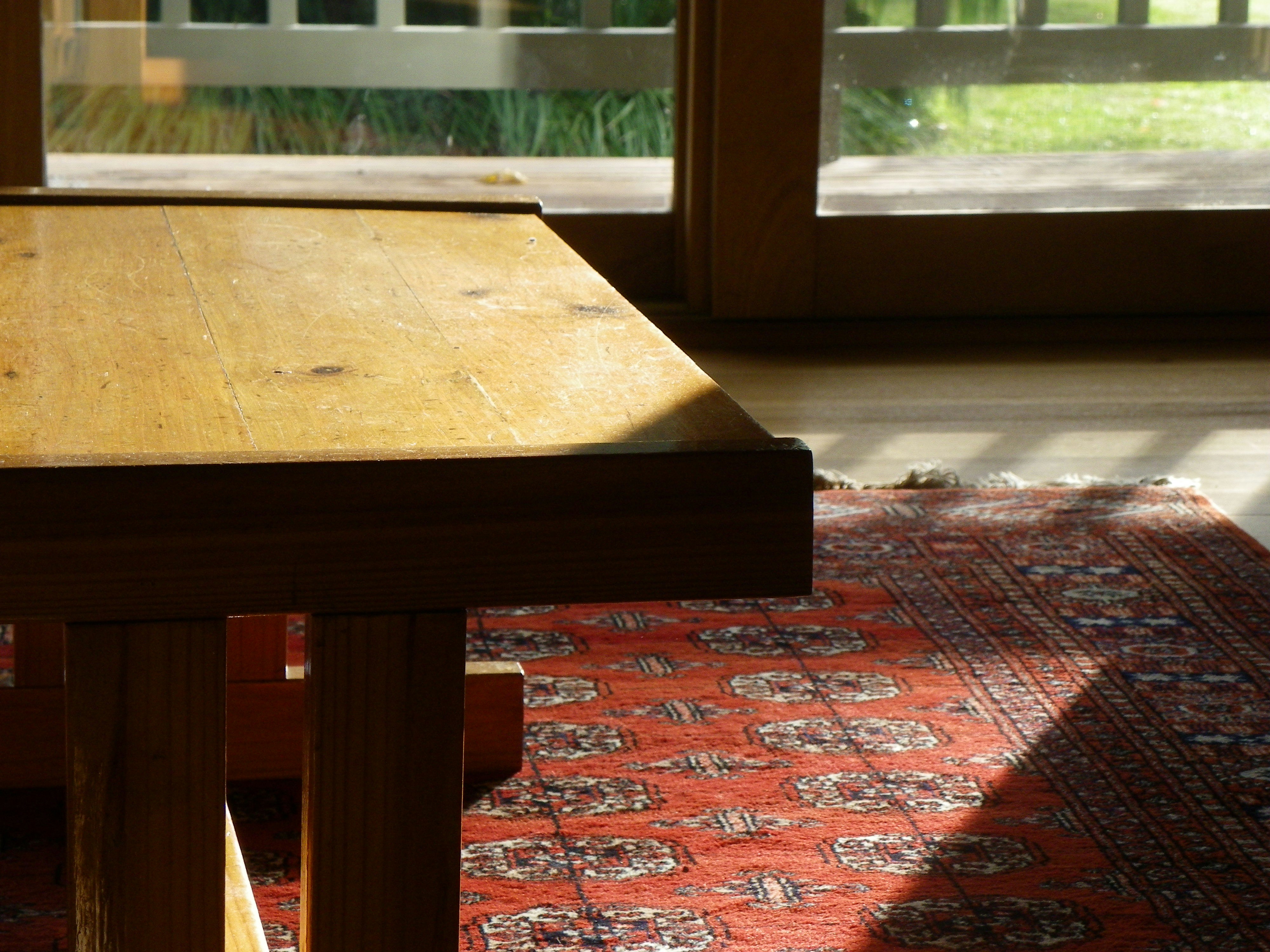 Sunlit wooden table sits on a crimson patterned rug by a window, with angular shadows filling the floor.