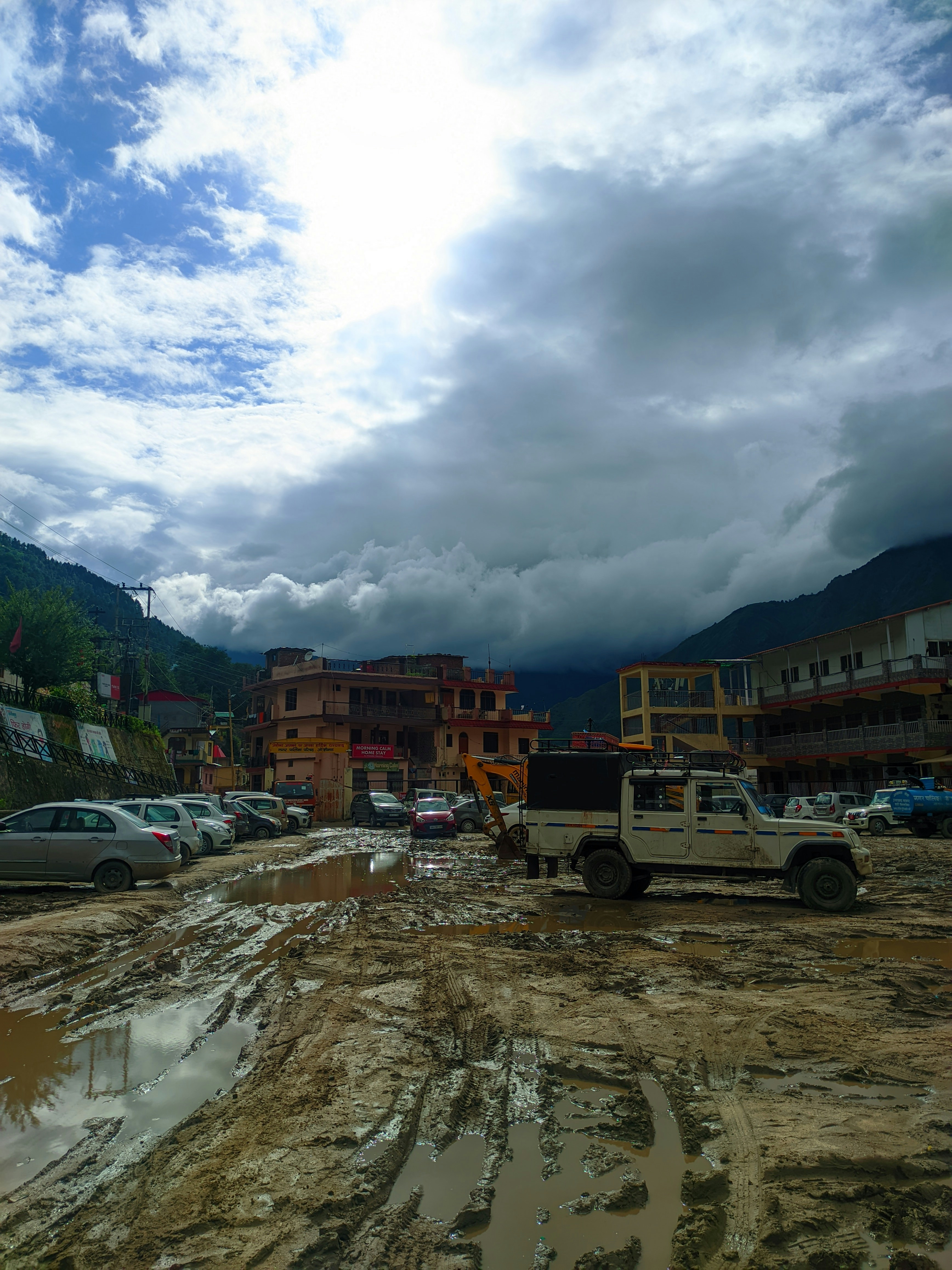 Mud-splashed street snakes through a small town, with parked cars and pastel buildings leading toward looming mountains. Puddles mirror the dramatic sky, lending a gritty, workaday mood.