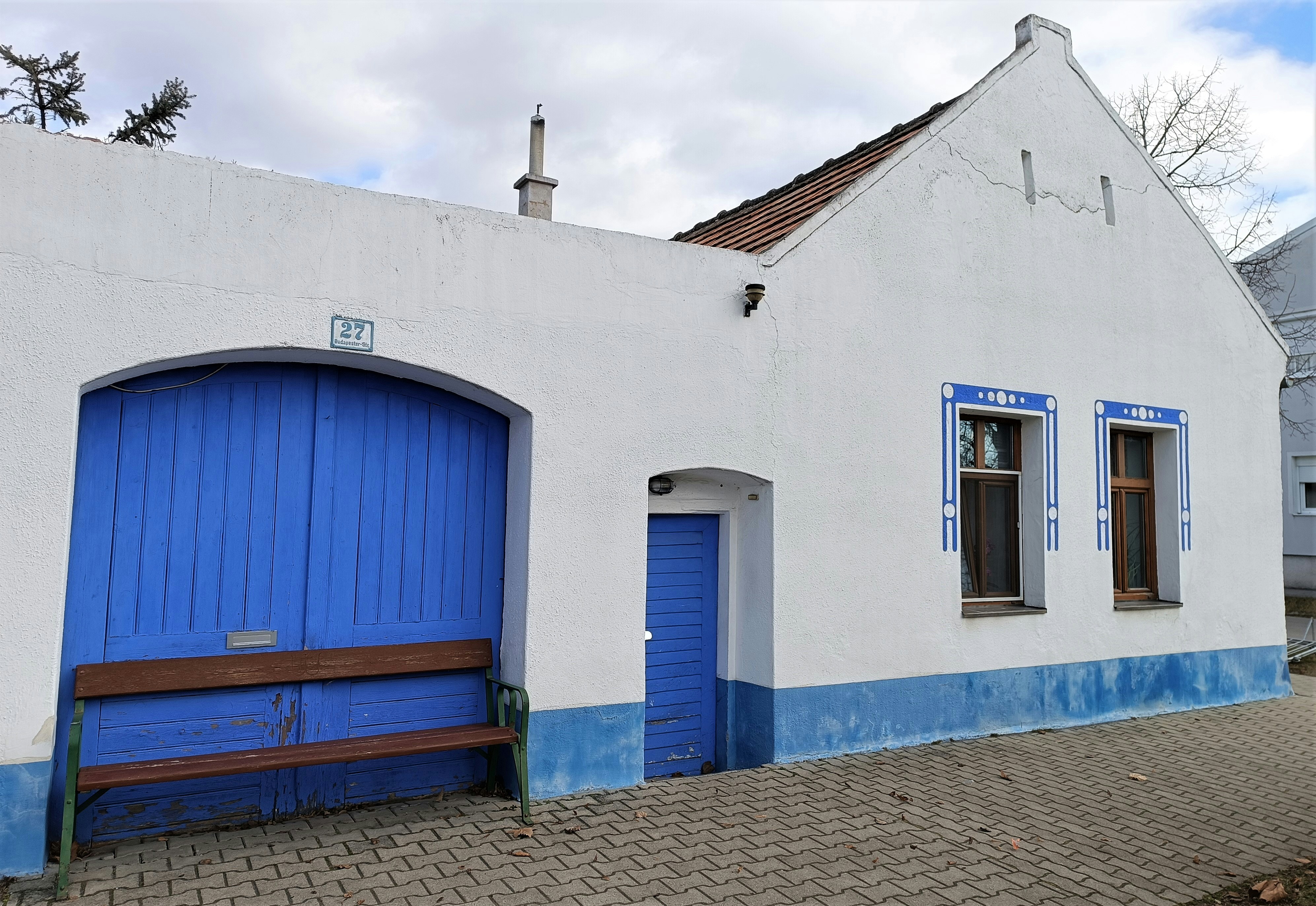 White-walled house with vivid blue doors and trim sits along a cobblestone street. A wooden bench rests beside the arched doorway, adding a rustic touch.