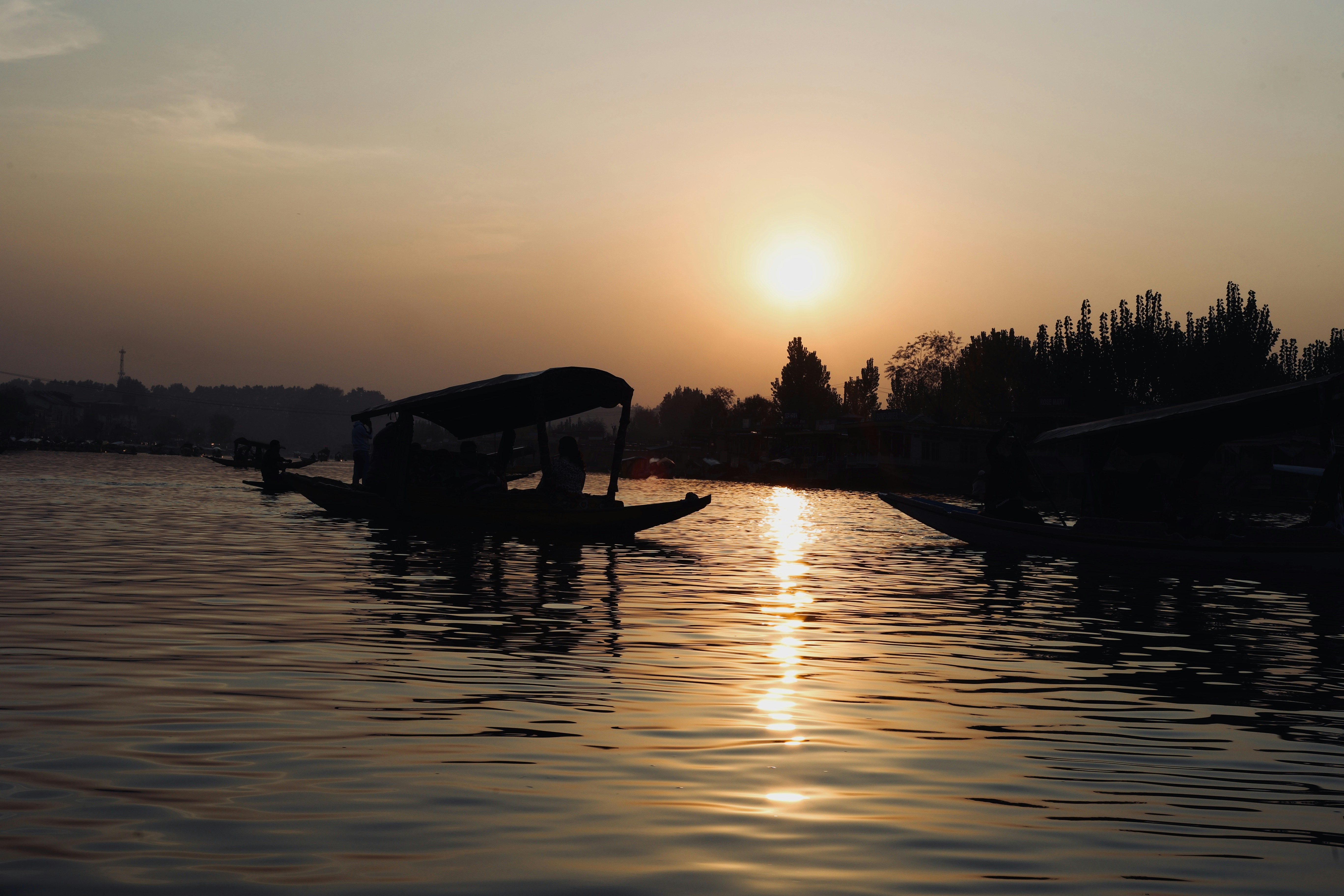 Silhouetted boats glide on a tranquil lake as the sun sets, casting warm reflections on the water.