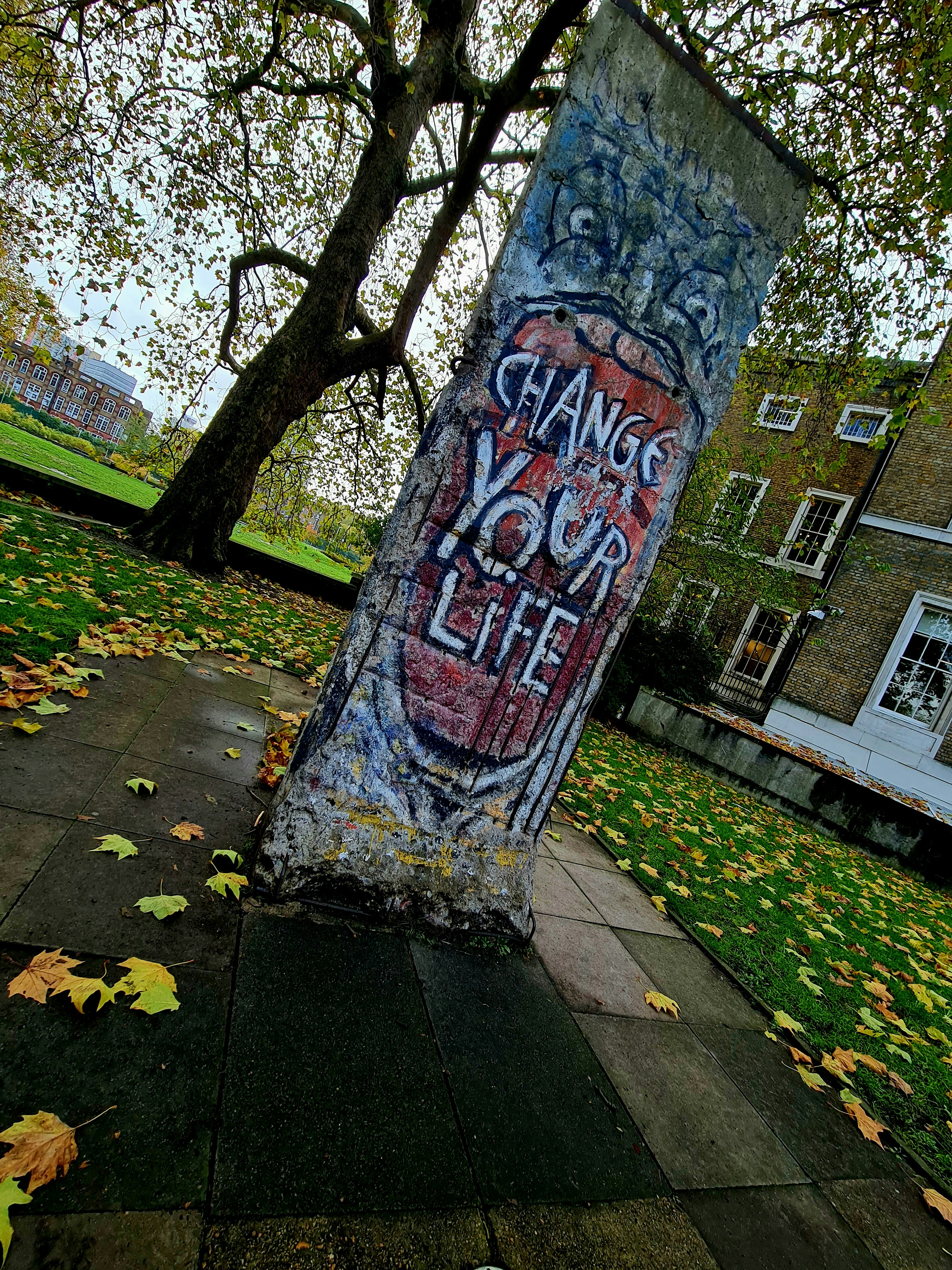 Graffiti-covered concrete pillar in a park, with autumn leaves on the ground and a brick building in the background.