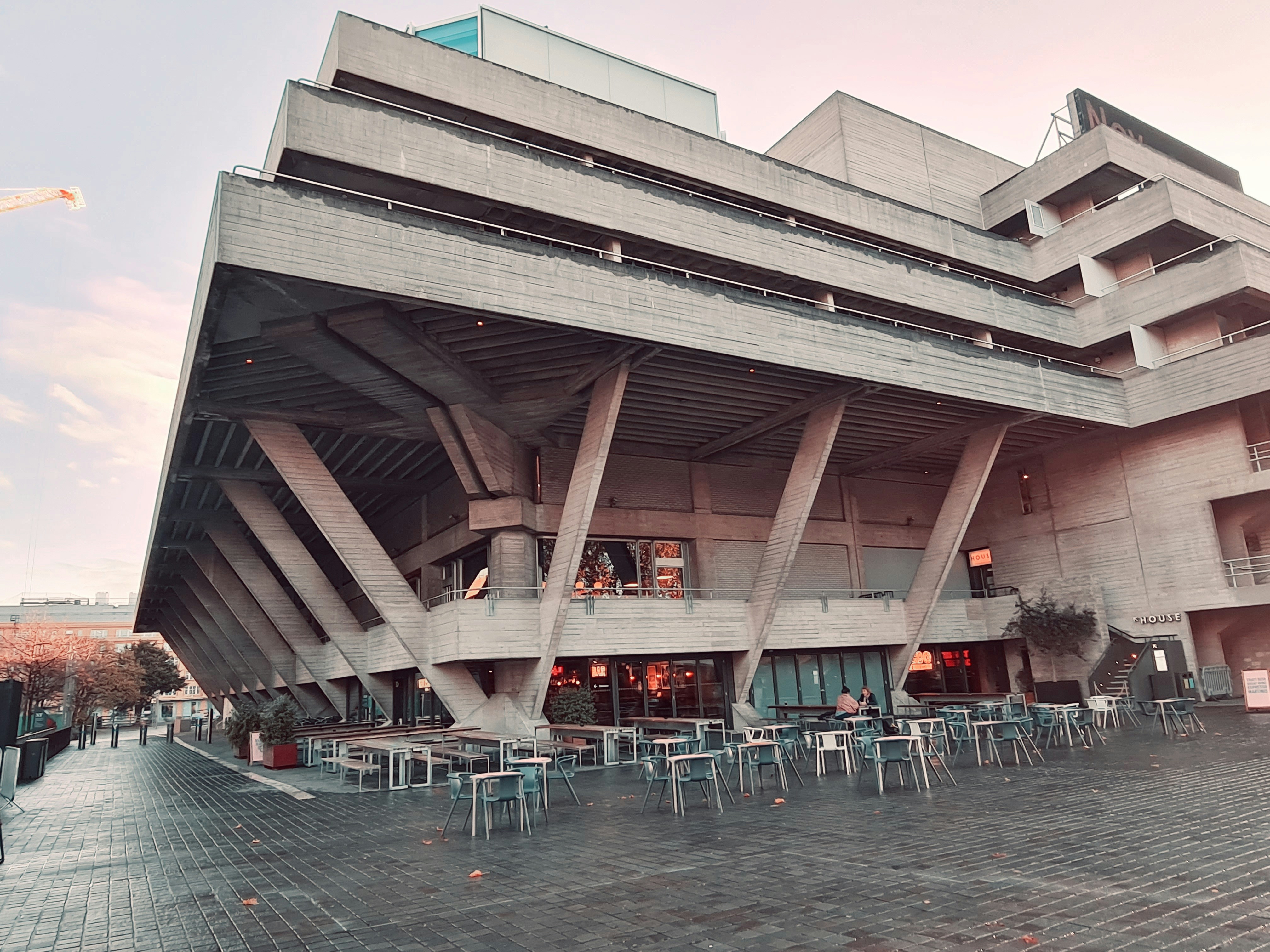 Concrete brutalist architecture with slanted supports looms over a sidewalk café. Pink dusk light spills across empty terrace chairs and the glass storefronts.