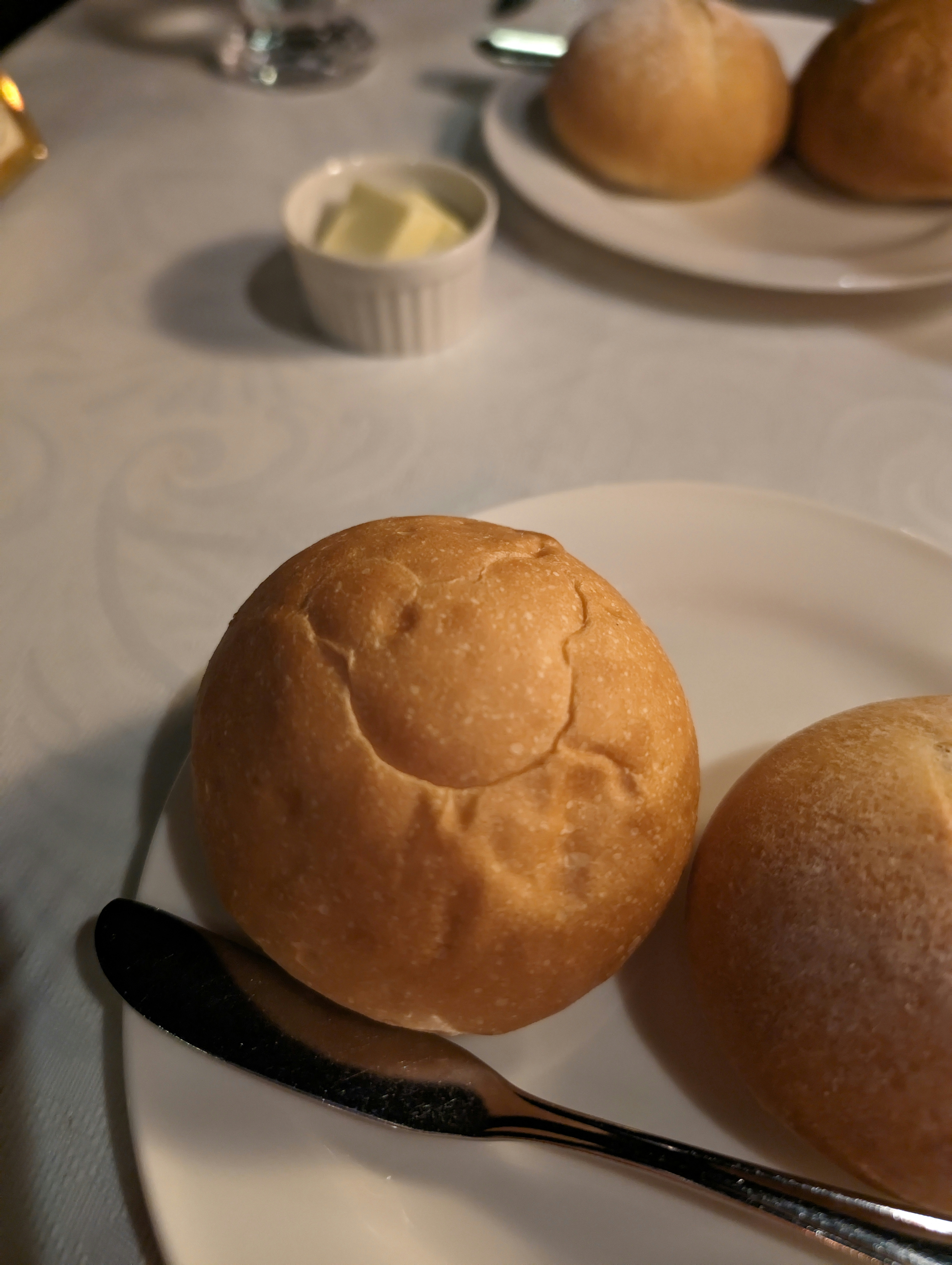 Close-up of golden dinner rolls on a white plate with a butter dish in the background. Soft restaurant lighting adds warm tones to the crust.