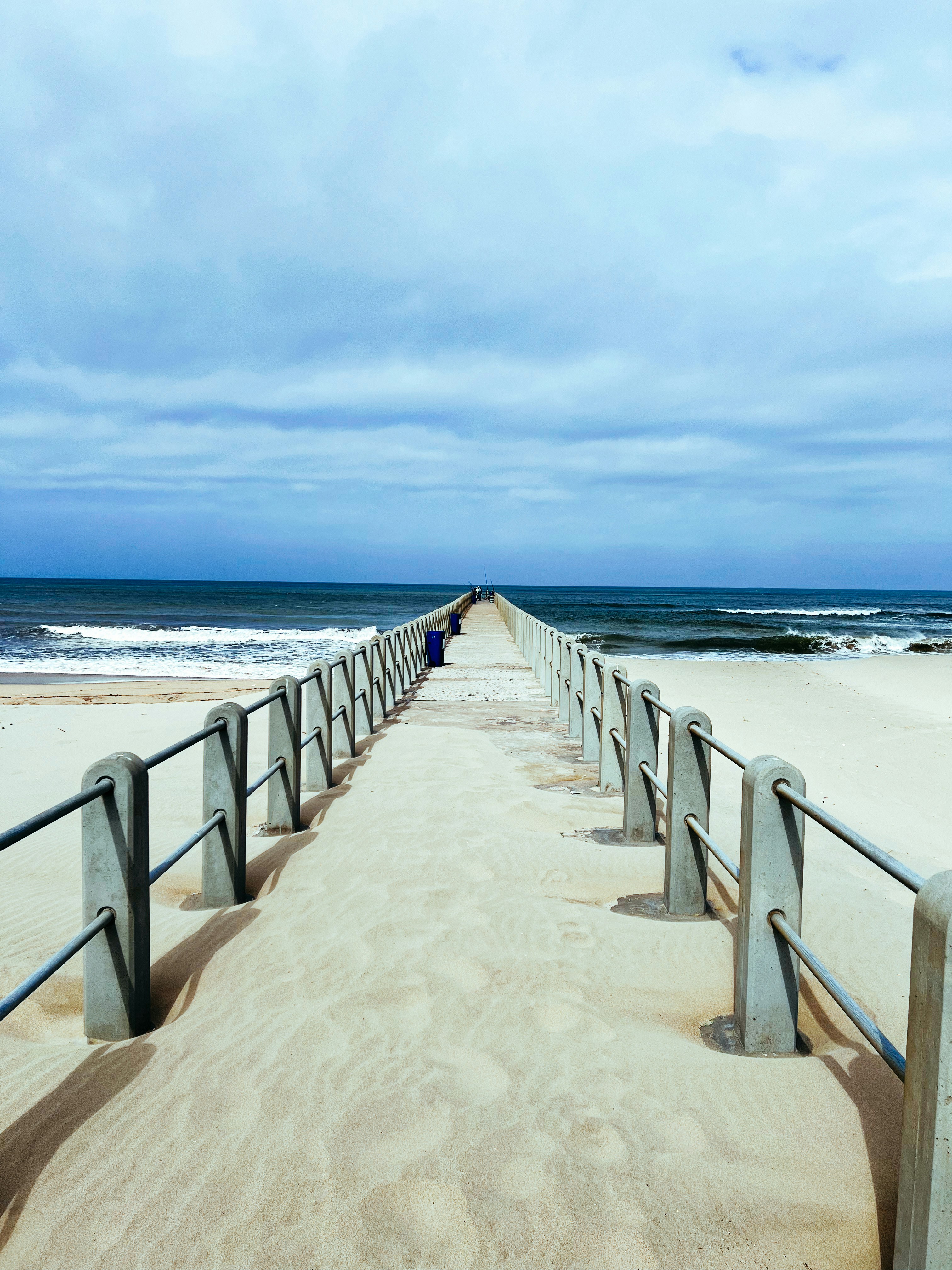A sandy pier stretches into the calm ocean, framed by gentle waves and a cloudy sky.