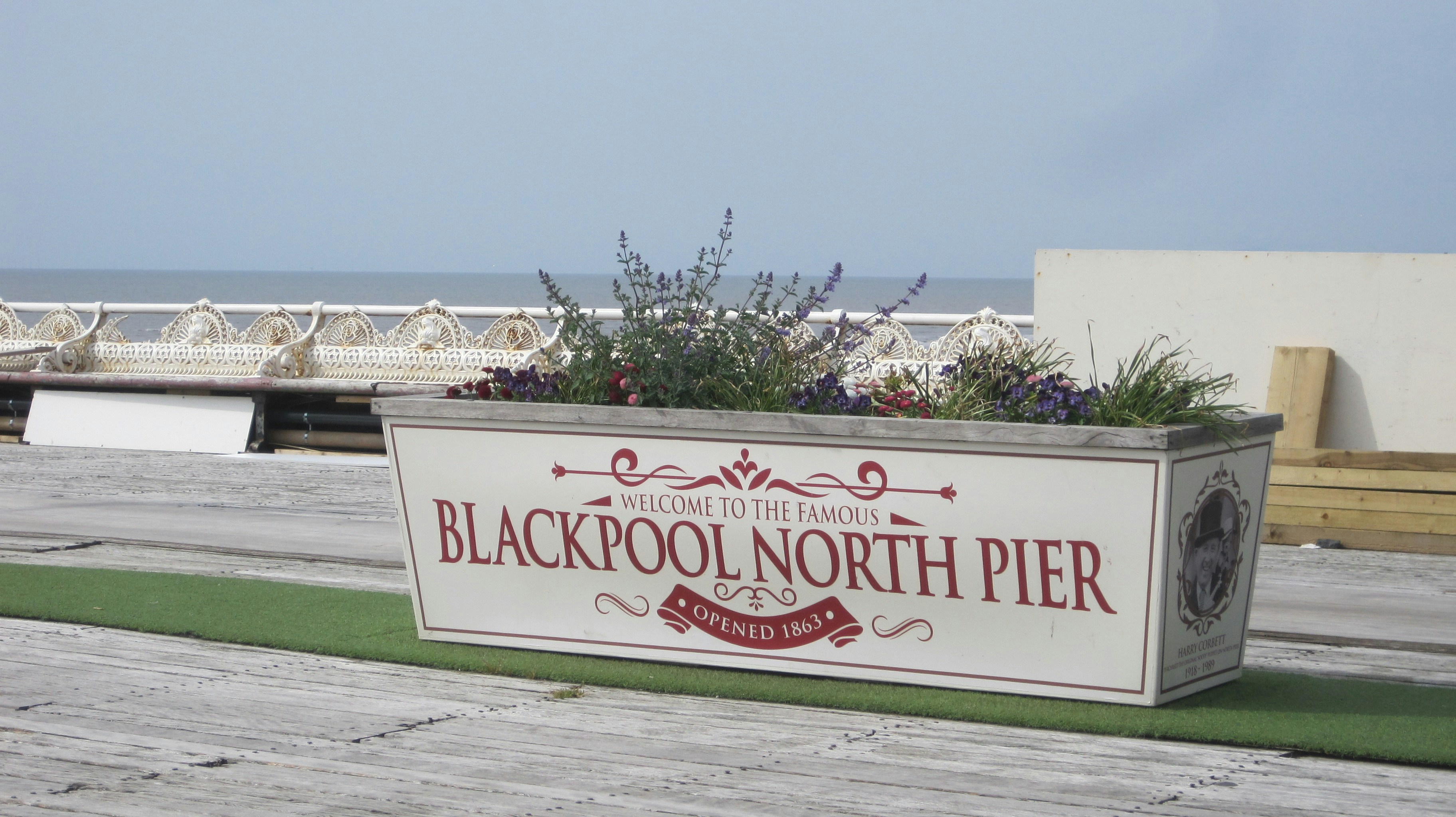Wooden planter labeled Blackpool North Pier brims with colorful flowers on a weathered boardwalk, with the sea and sky in the background.