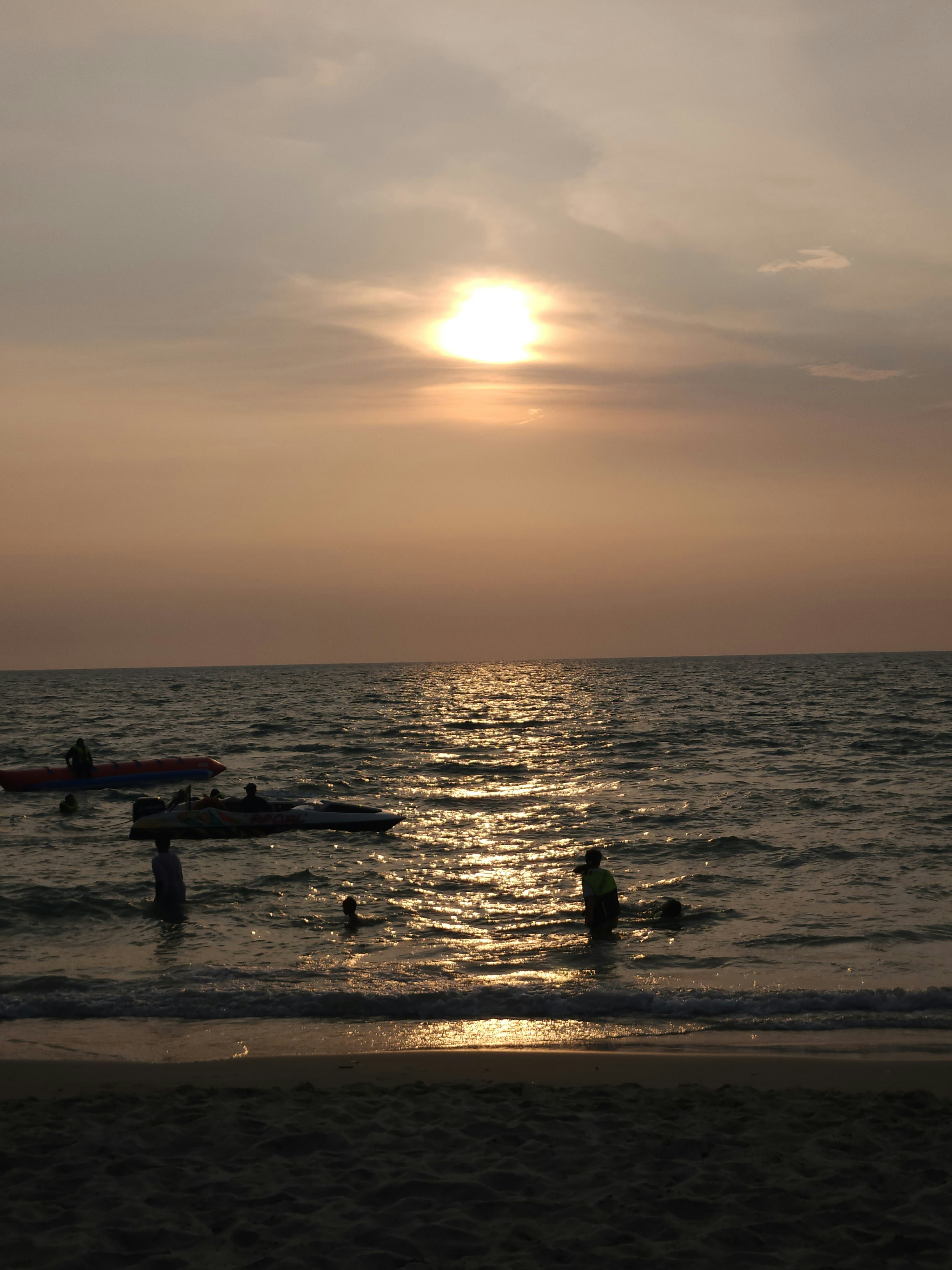 Sunset over a calm sea with silhouettes of swimmers near the shore and distant boats on the horizon. The warm, diffused light softens the coastline as the sky glows with amber tones.