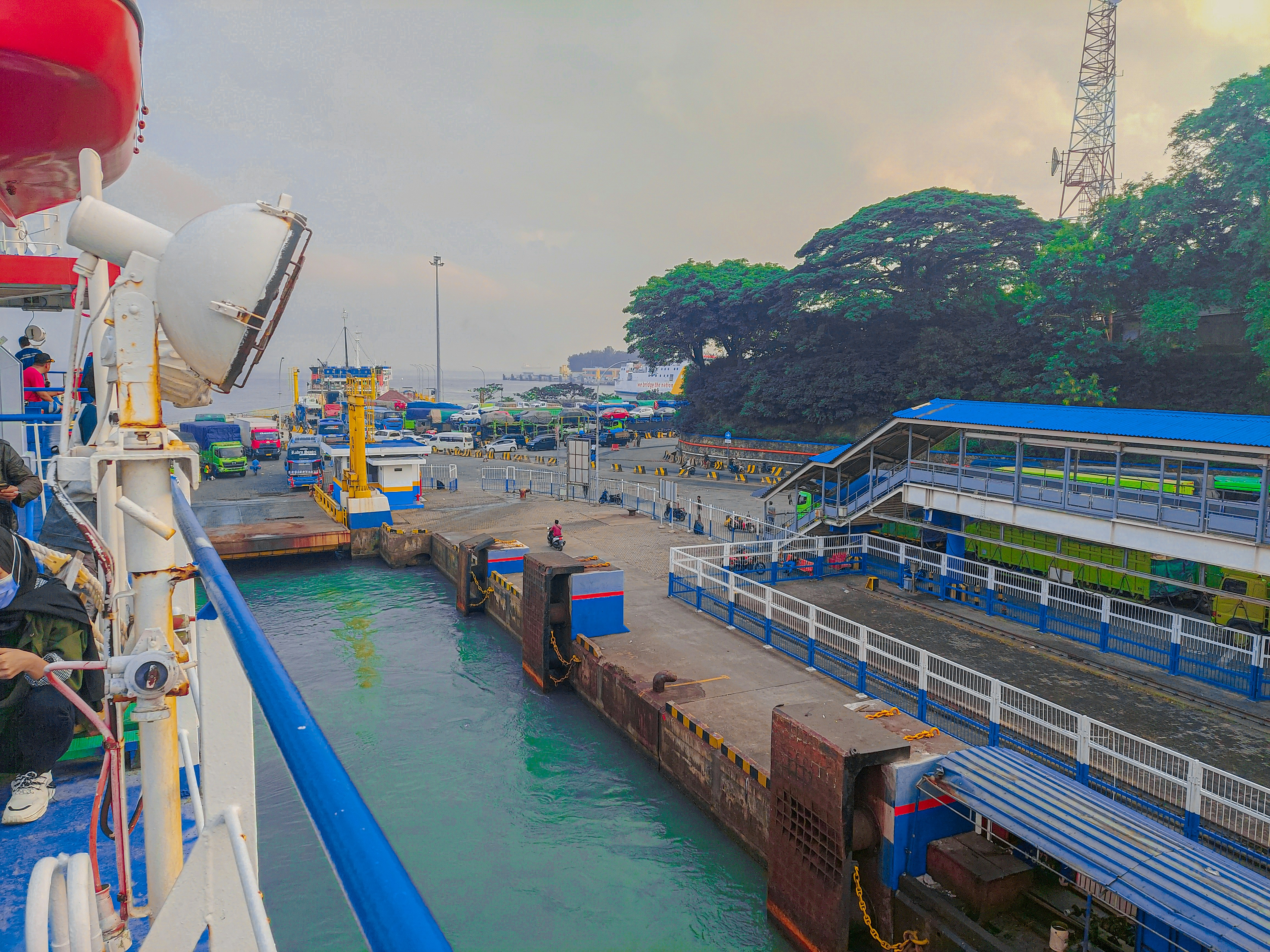 Docked ferry alongside bustling port with vivid blue accents and distant greenery at dawn.