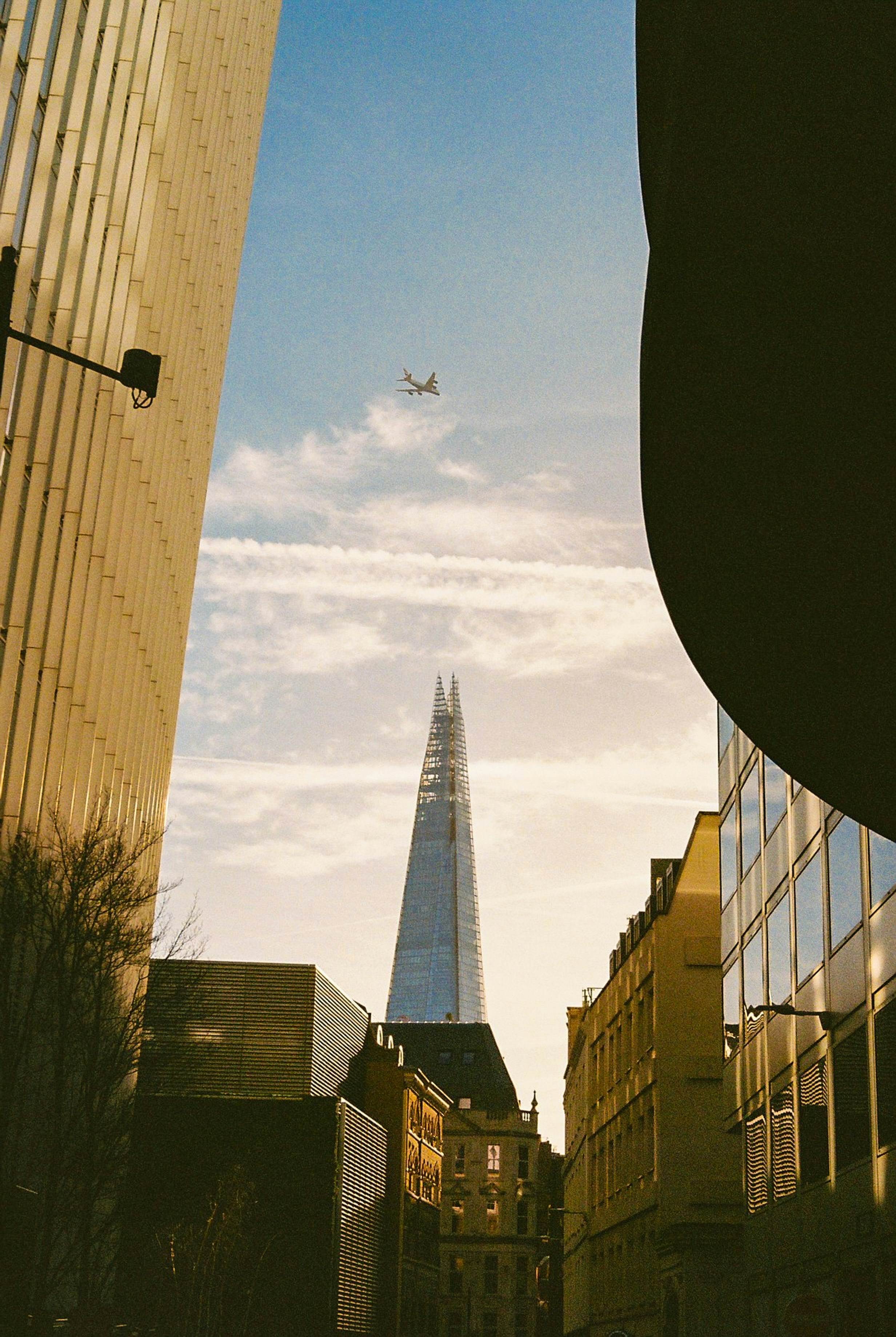 Urban canyons frame a direct view toward The Shard as the street narrows into the distance. A small airplane crosses the pale blue sky above.